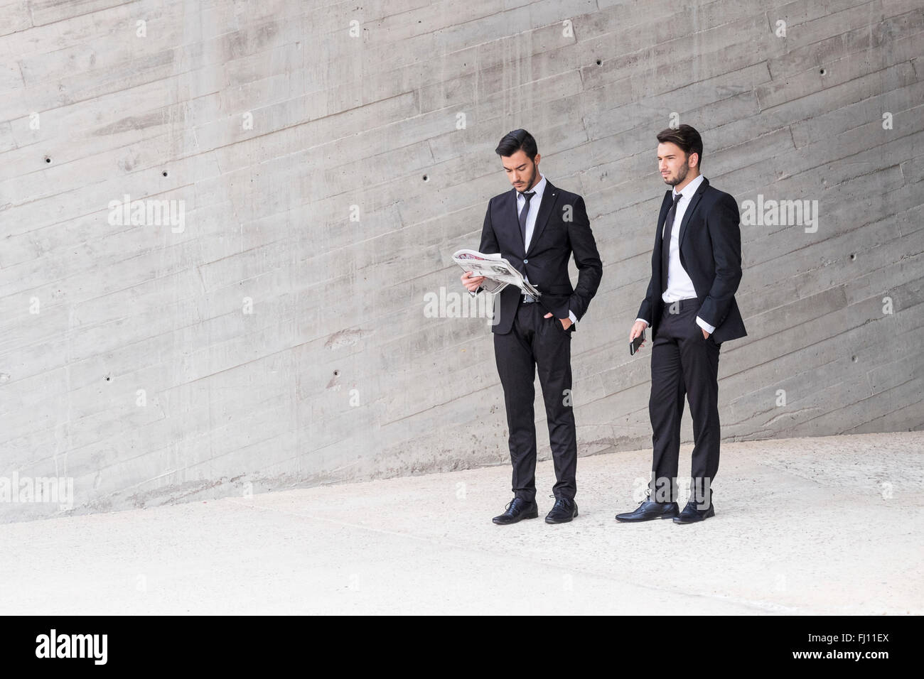 Two businessmen wearing black suits standing in front of concrete wall ...