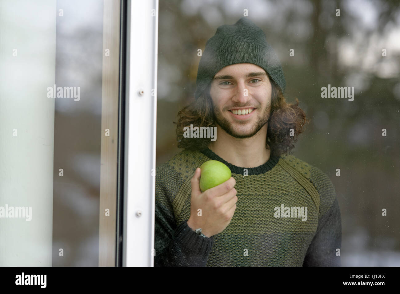 Portrait of smiling young man with green apple standing behind window ...