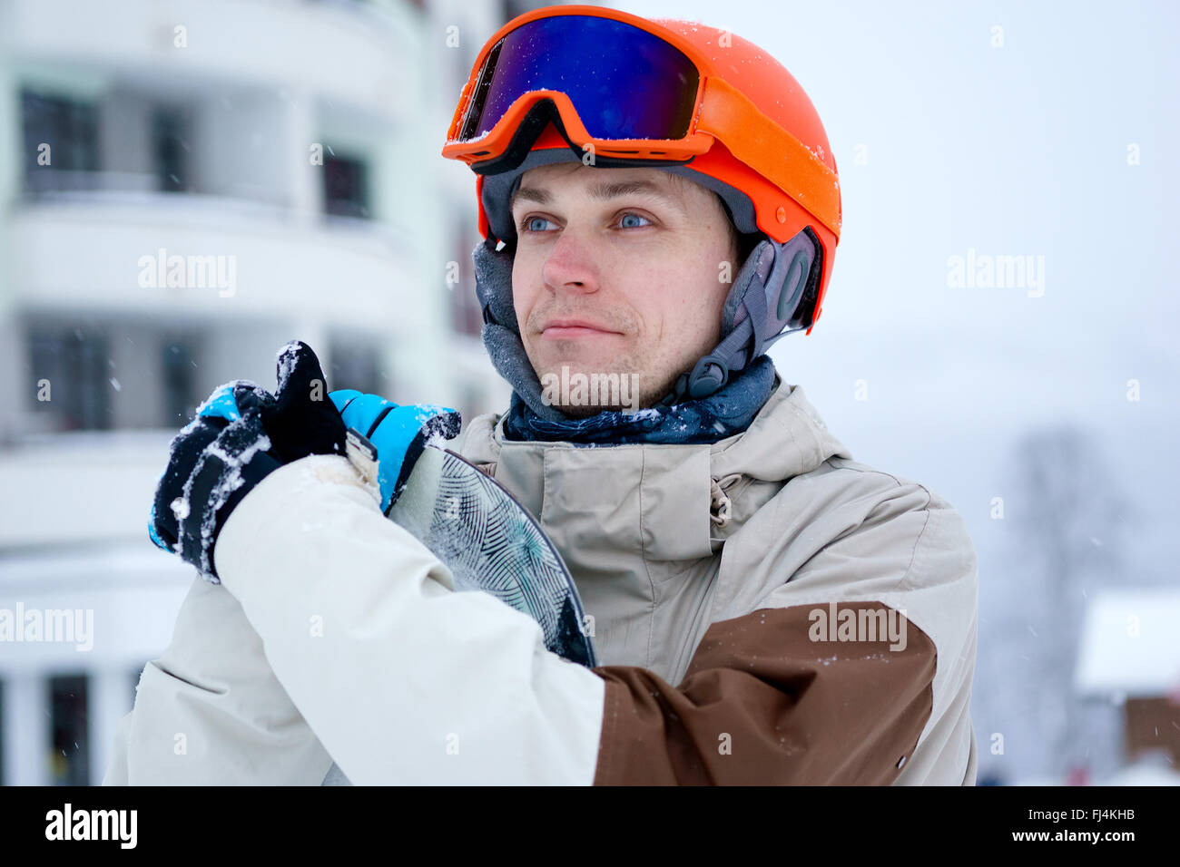 Man snowboarder wearing orange helmet, grey jacket, black and blue gloves standing with ...