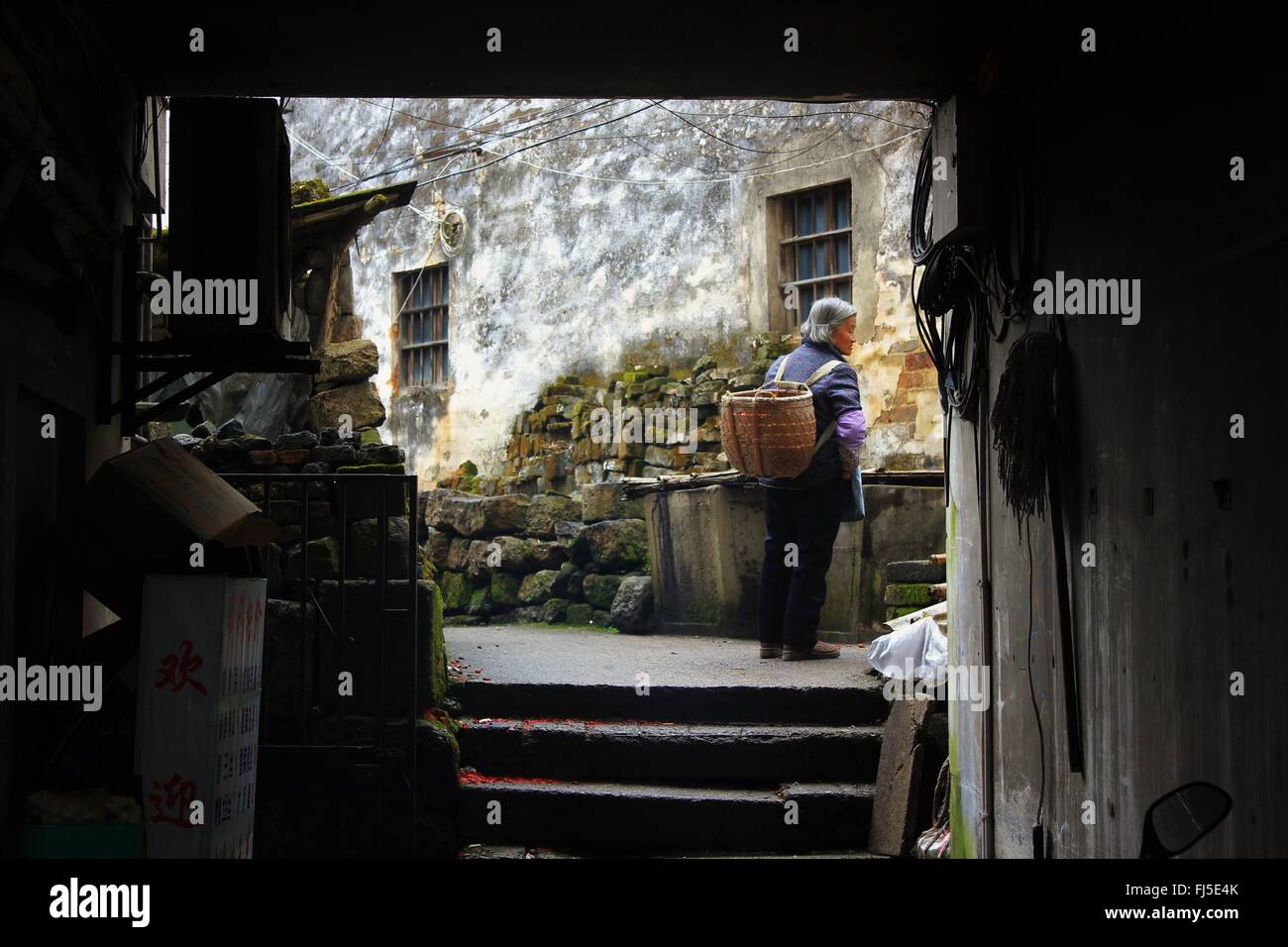 Elderly Chinese lady carrying a basket standing in a rustic alleyway in ...
