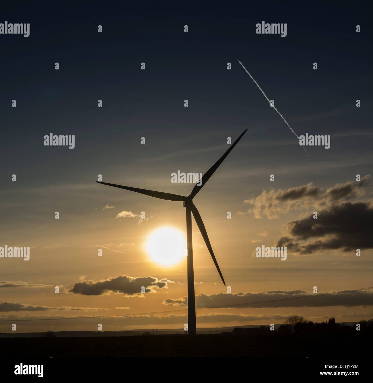 Wind turbine generator against sunset with airplane con trail overhead ...