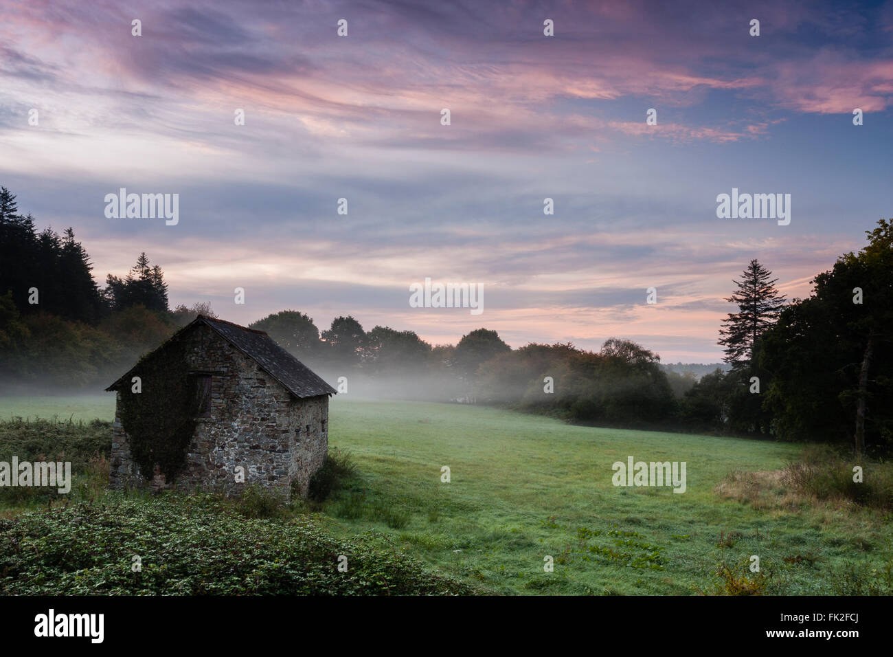A small stone hut stands at a grass field as the morning mist rises ...