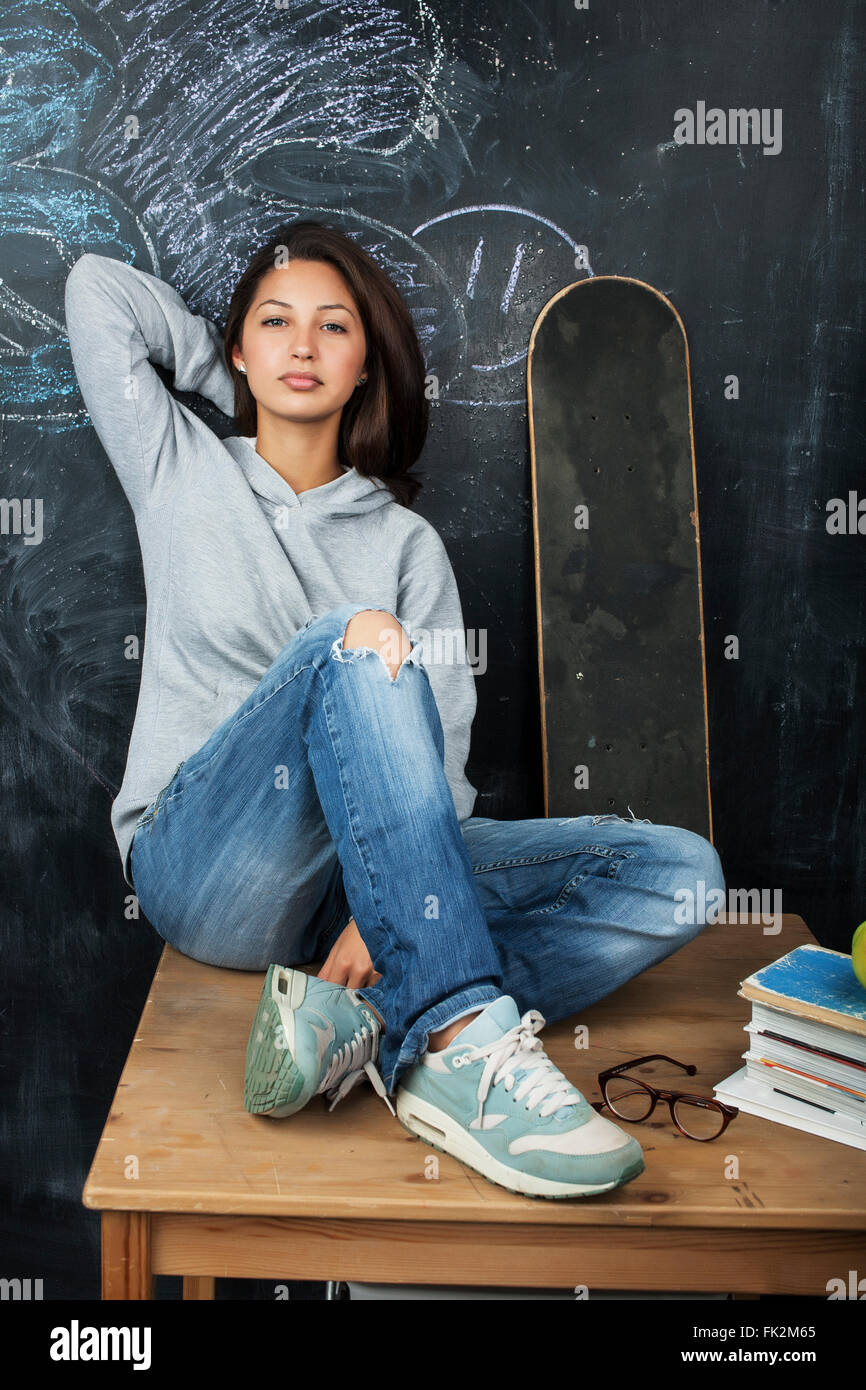 young cute teenage girl in classroom at blackboard seating on table ...