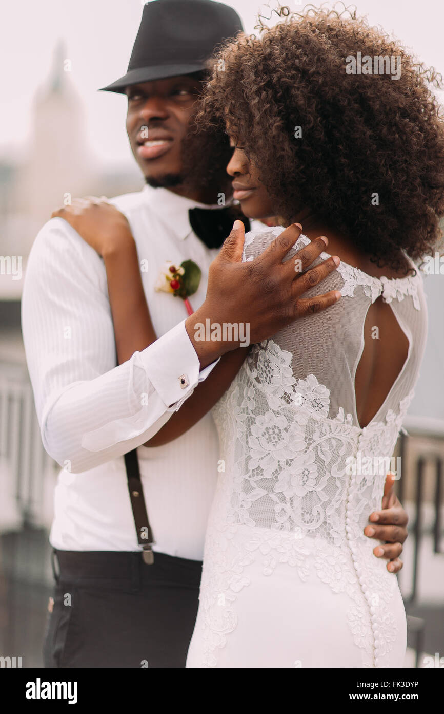 Happy stylish african groom in hat and suspenders holding his pretty ...