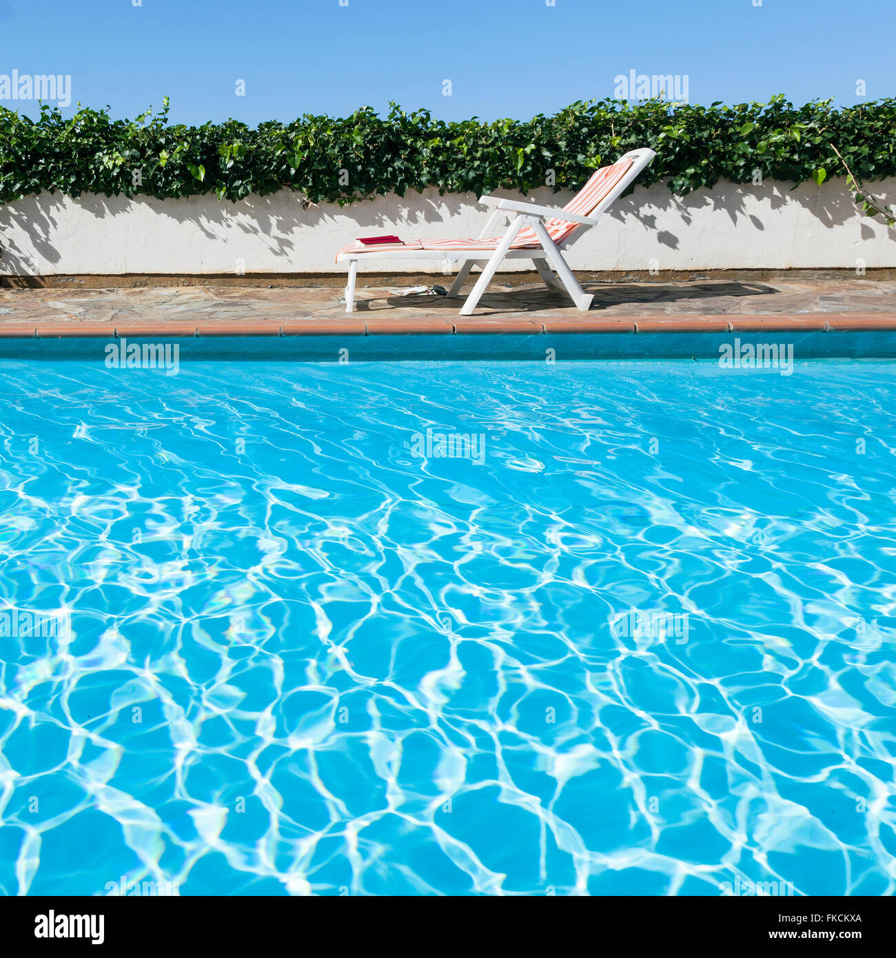 ripples in swimming pool with ivy on wall and blue sky and deck chair ...