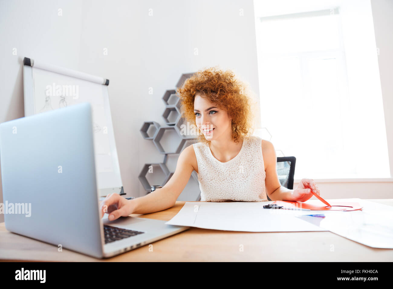Cheerful curly redhead young woman cutting color paper and using laptop ...