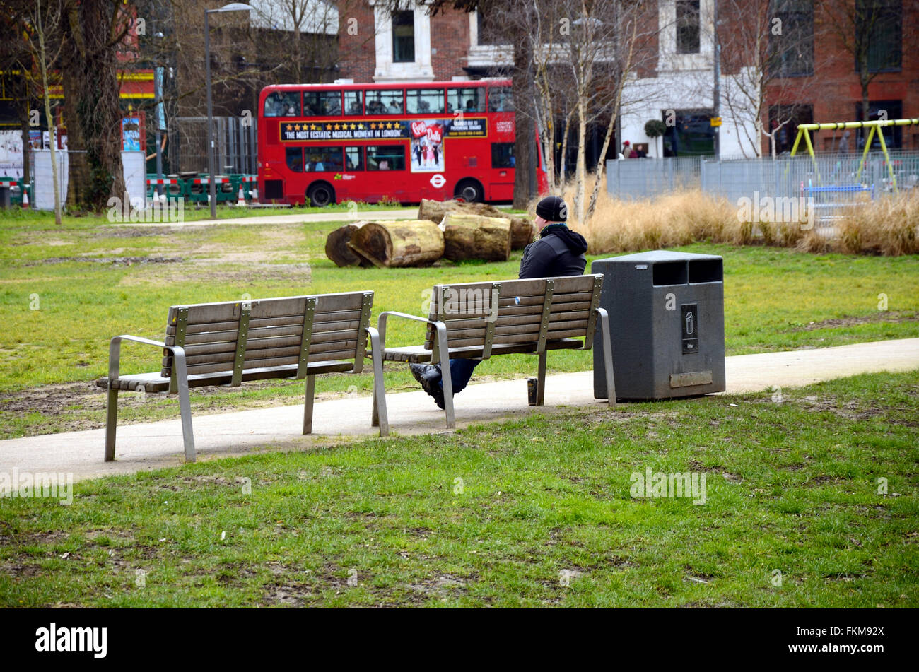 A man sits on a park bench in the middle of Shepherds Bush Common Stock ...