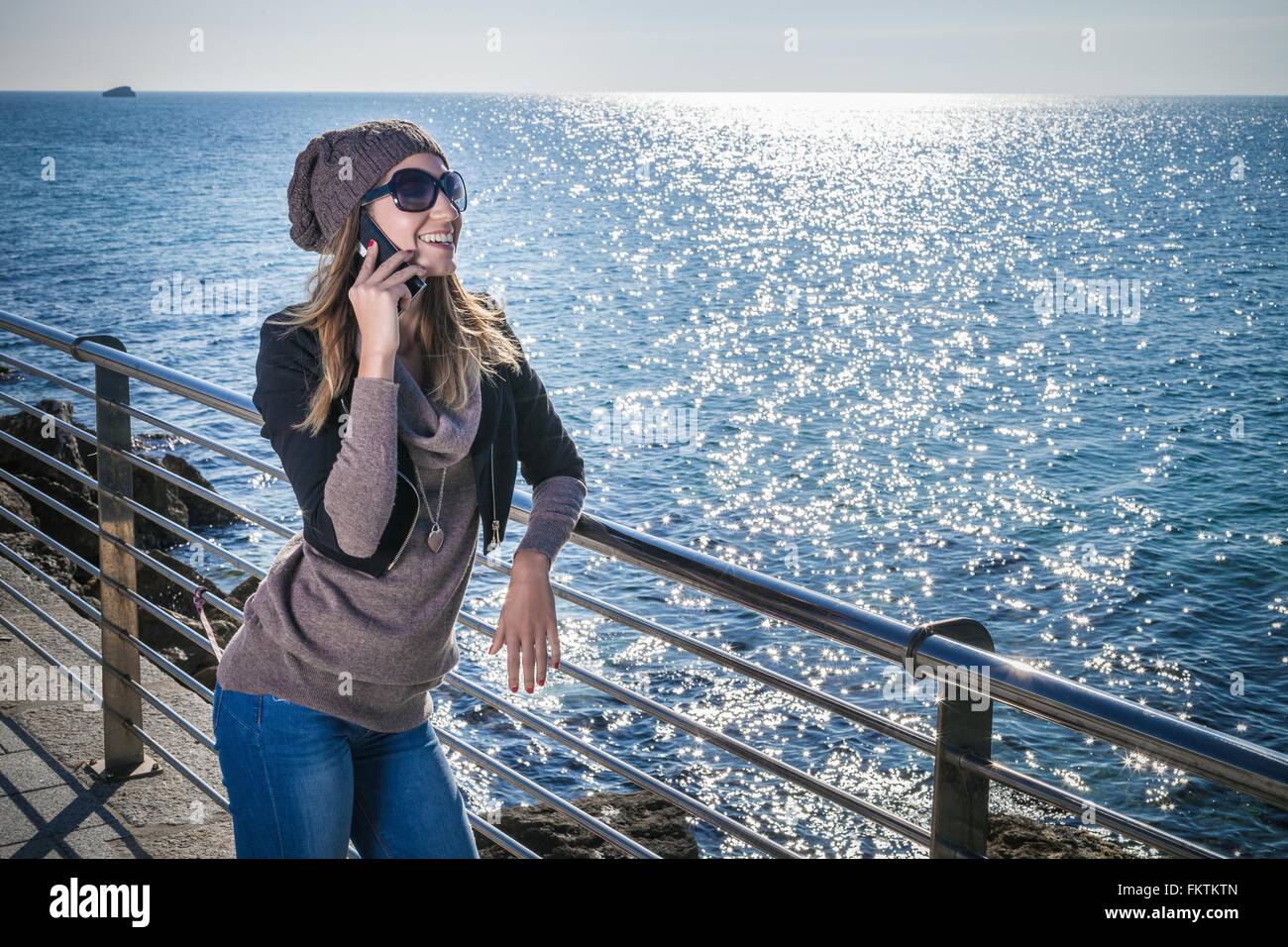 Young woman leaning against railings by ocean using smartphone looking ...