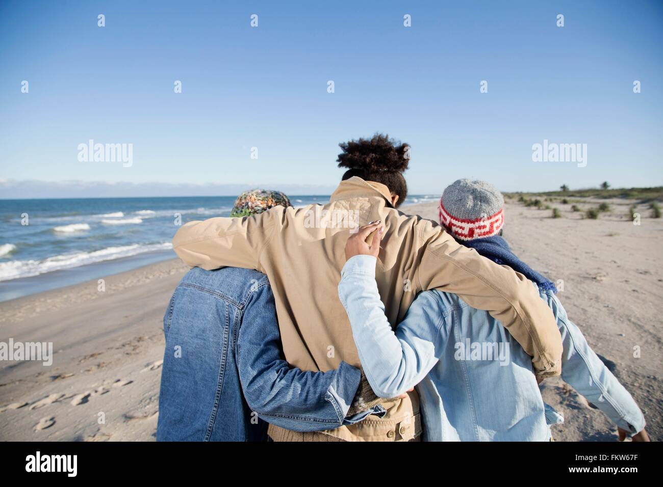 Three friends walking along beach, arms around each other, rear view ...