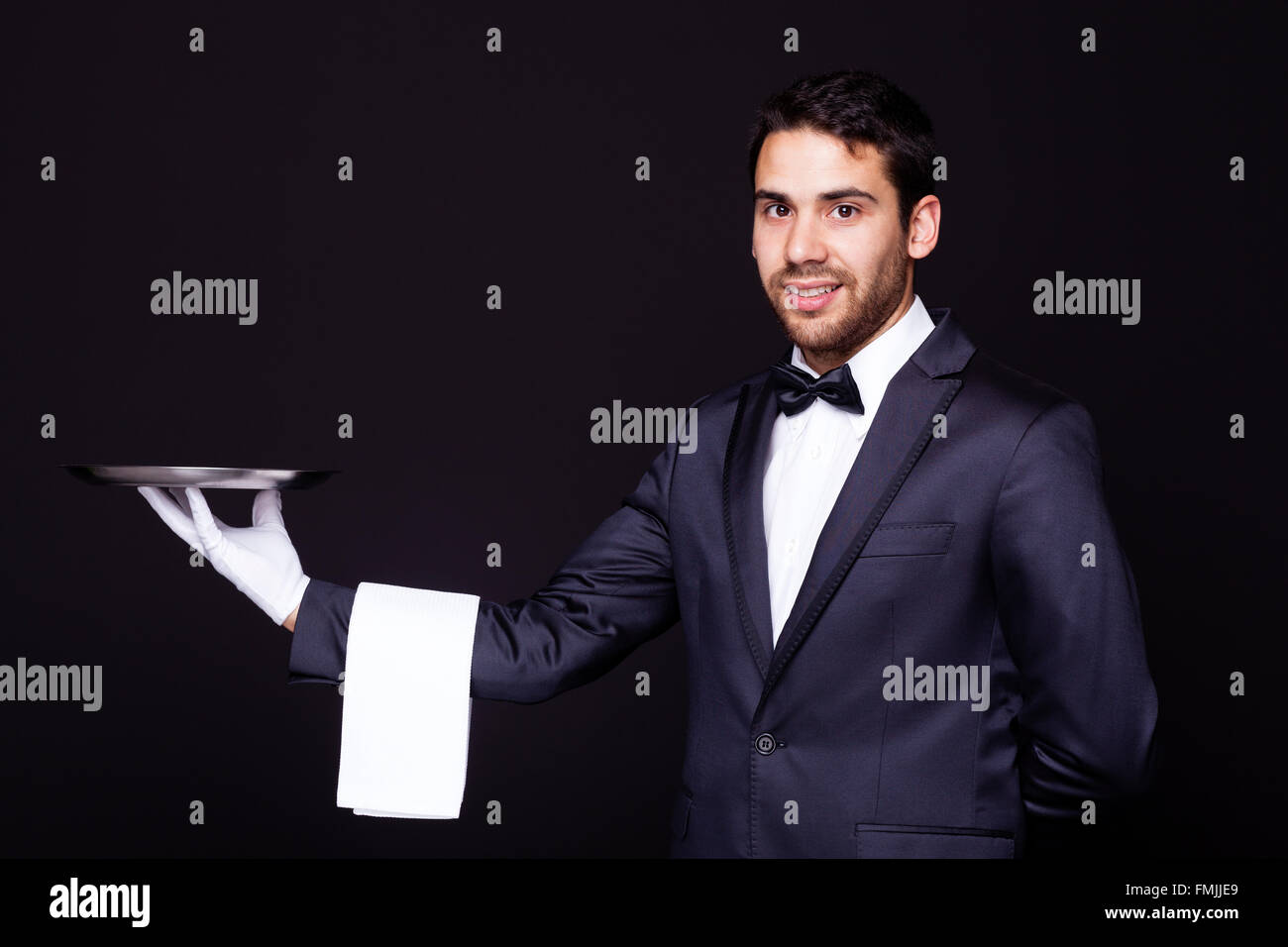 Portrait of a smiling waiter holding an empty silver tray against dark ...