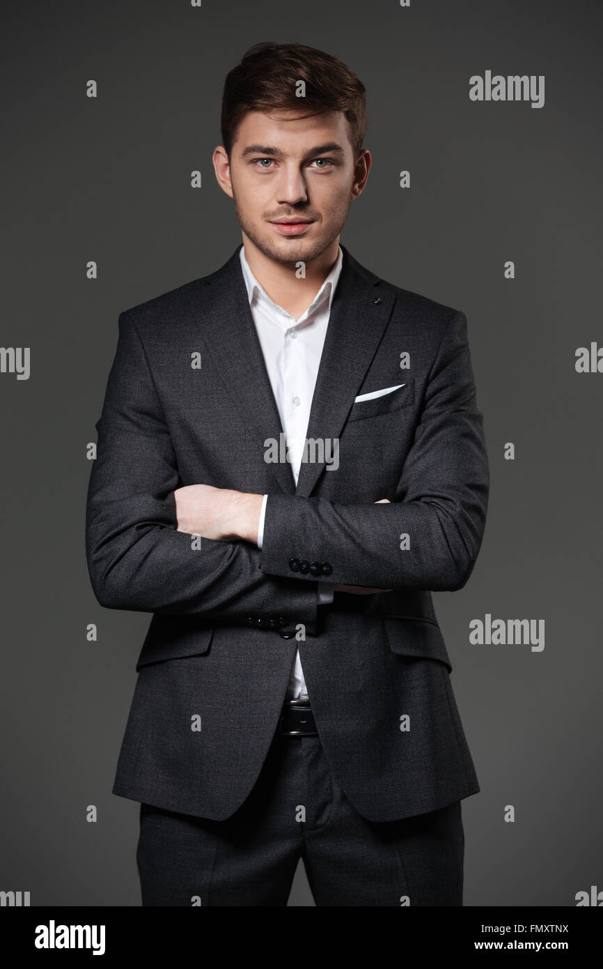 Portrait of handsome confident young businessman in black suit standing with arms crossed over ...