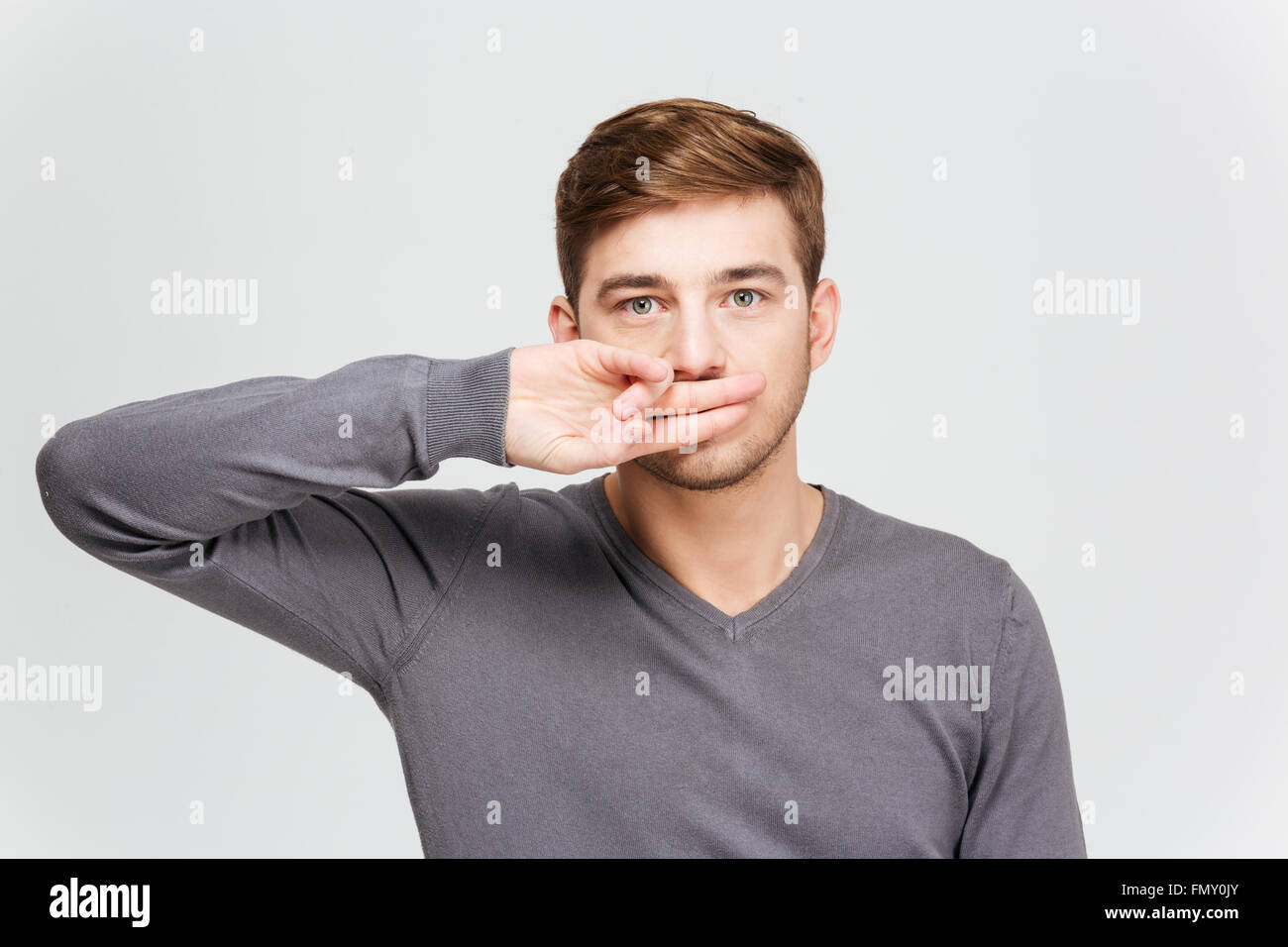 Serious handsome young man in grey pullover covered mouth by fingers ...