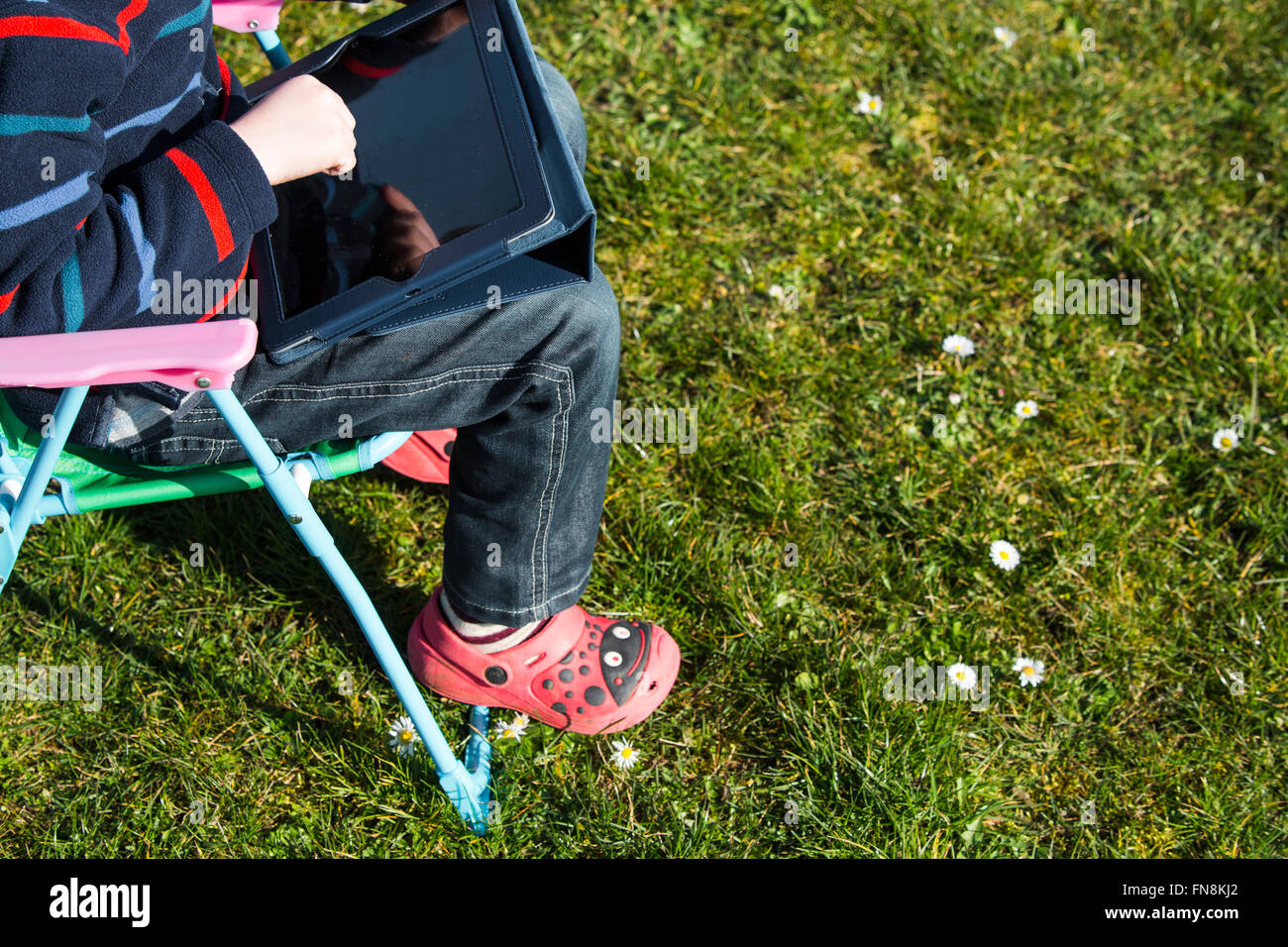 A young child using an ipad while sat outside on a small deck chair ...