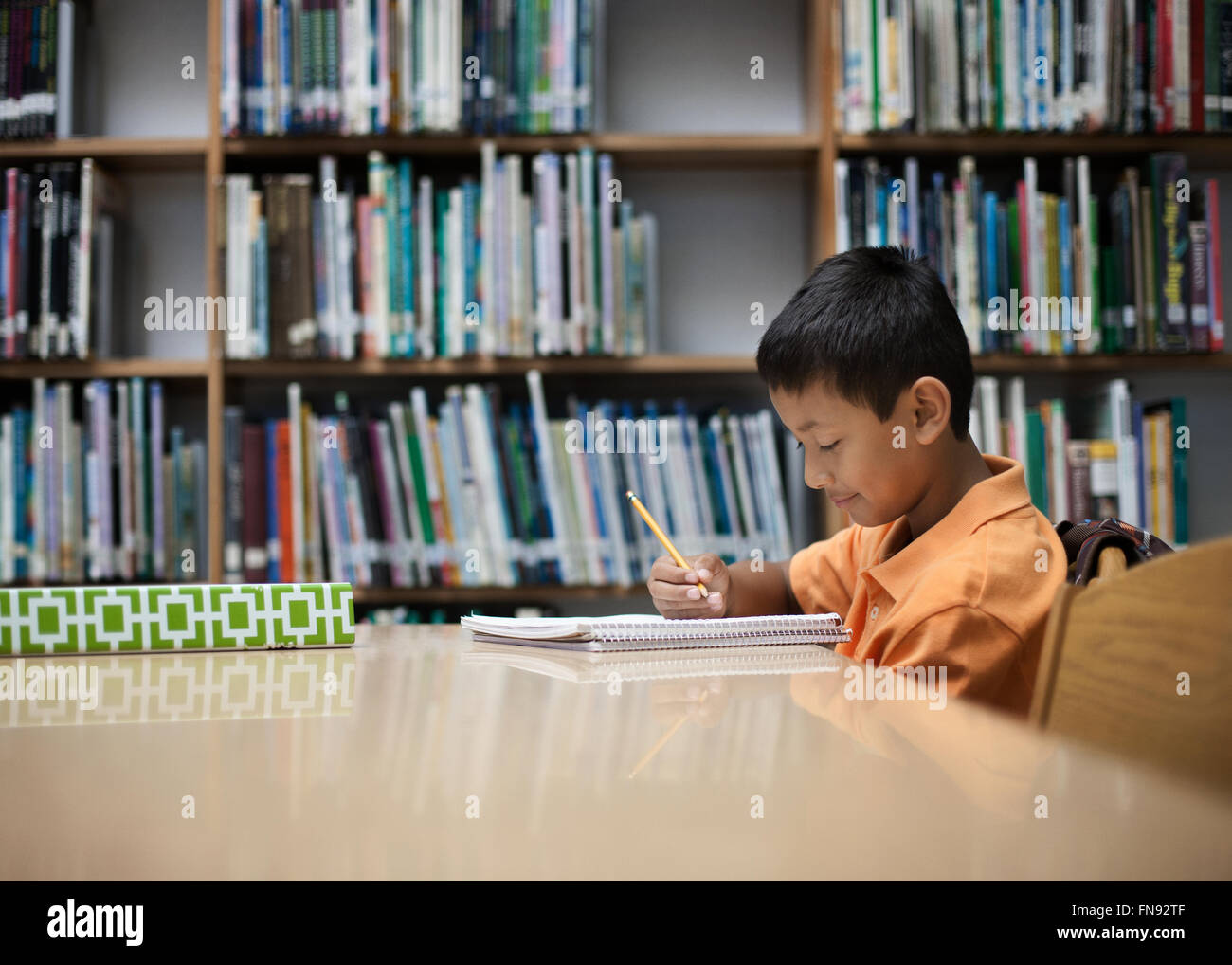 A boy sitting at table in a school library, using a pencil, studying ...