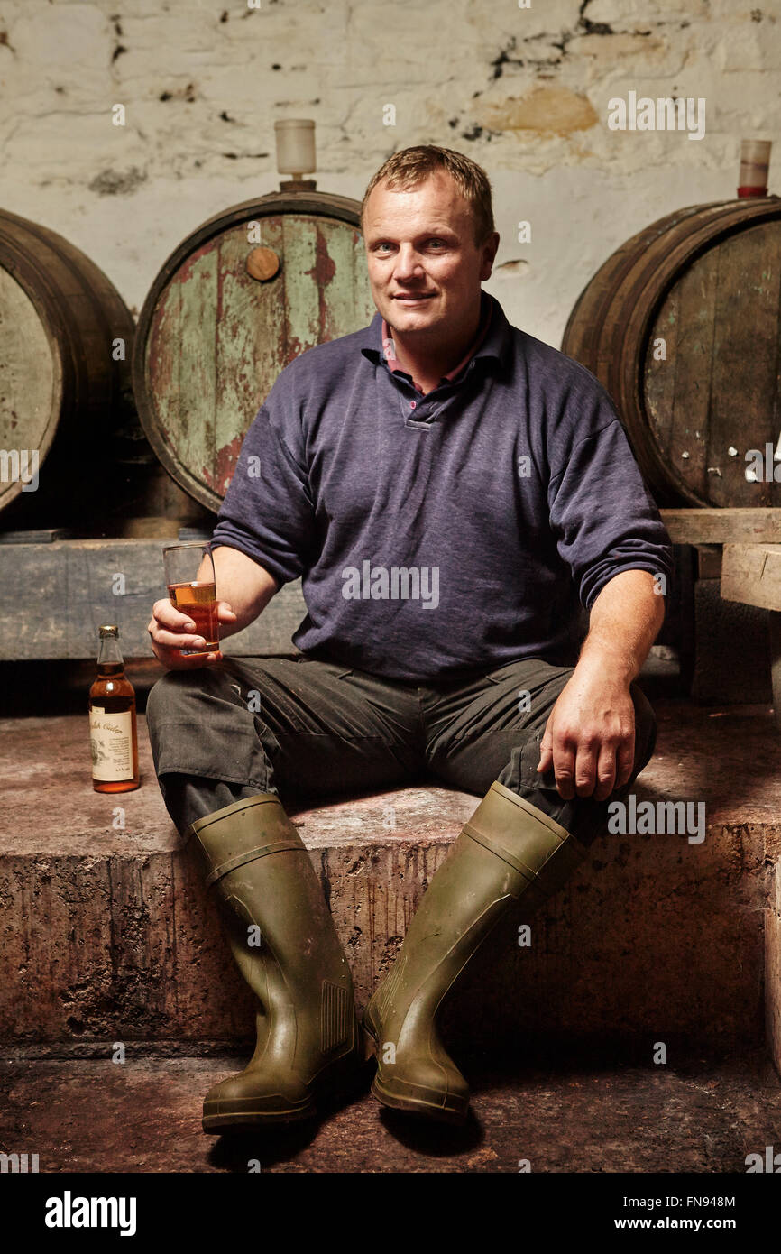 A man sitting among oak barrels at a cider makers, raising a glass and ...