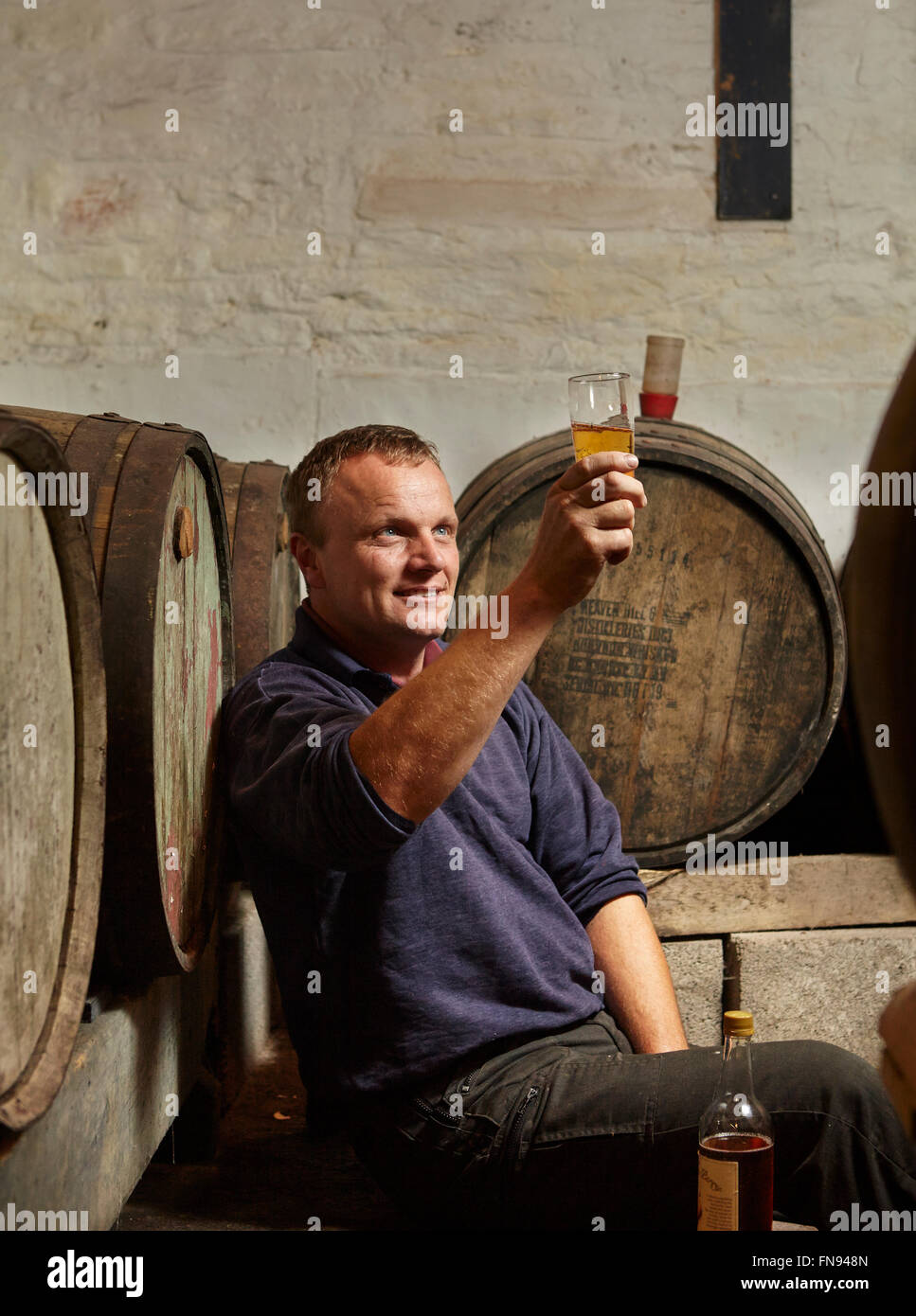 A man sitting among oak barrels at a cider makers, raising a glass and ...
