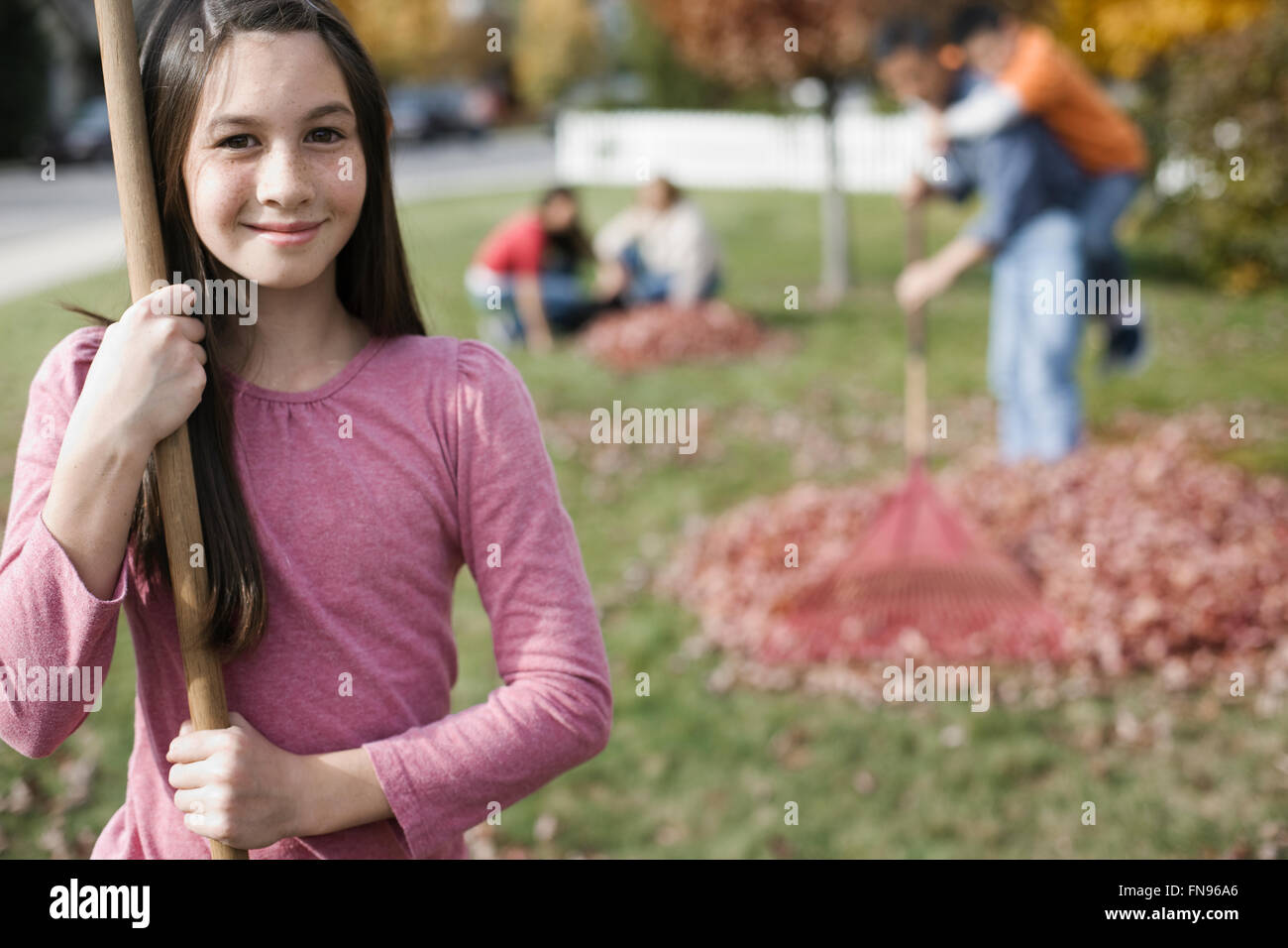 A girl holding a leaf rake and people raking fallen autumn leaves into ...