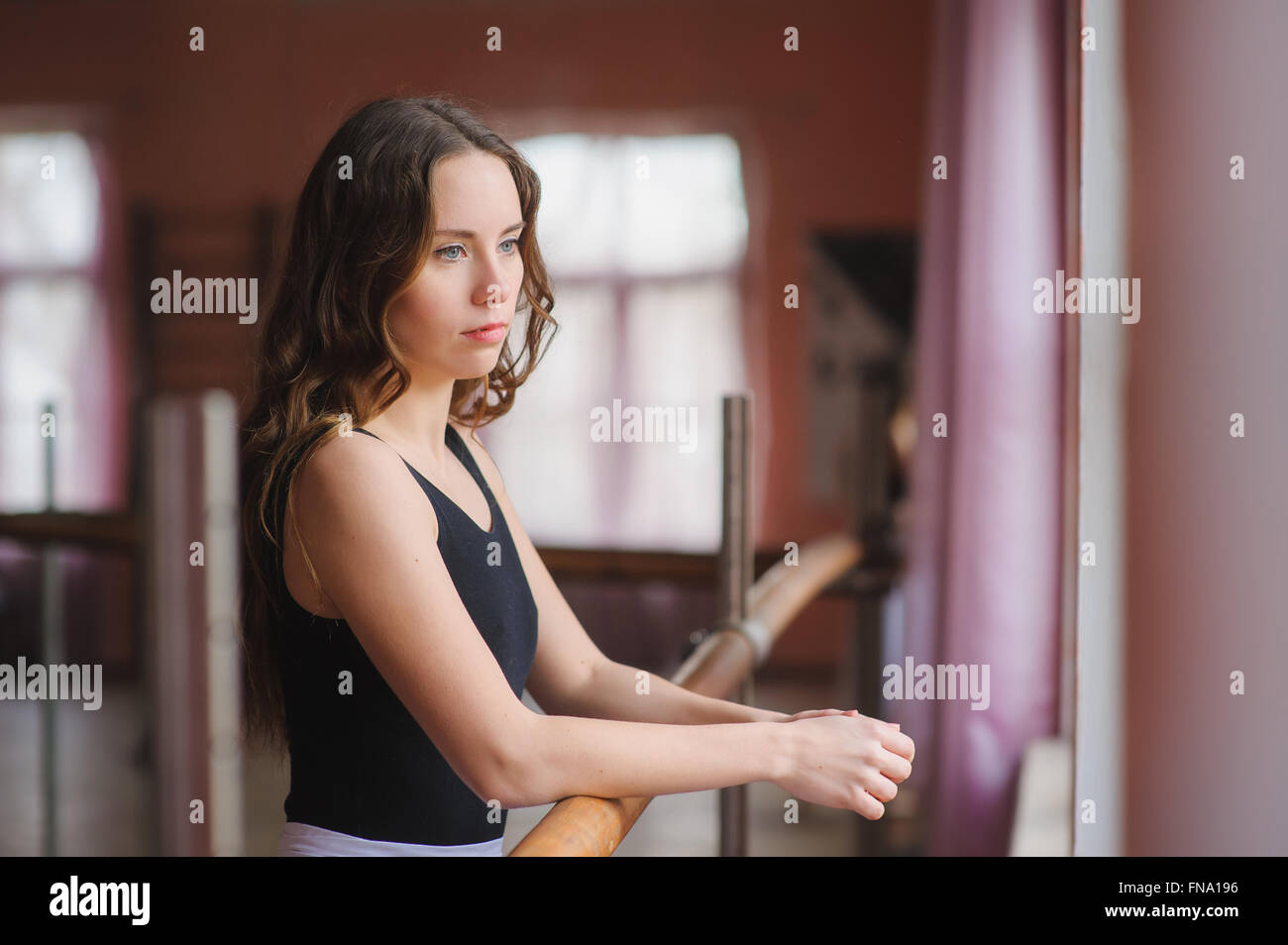 Thoughtful girl dancer. Beautiful young ballerina holding hand on ballet barre while looking ...