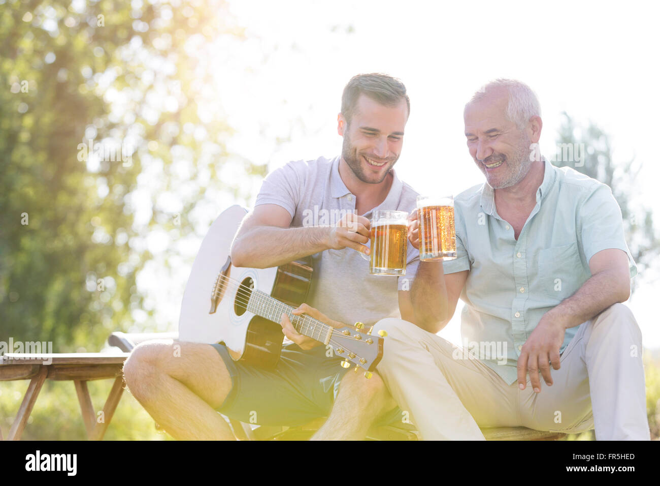 Father And Adult Son Toasting Beer Mugs And Playing Guitar Outdoors