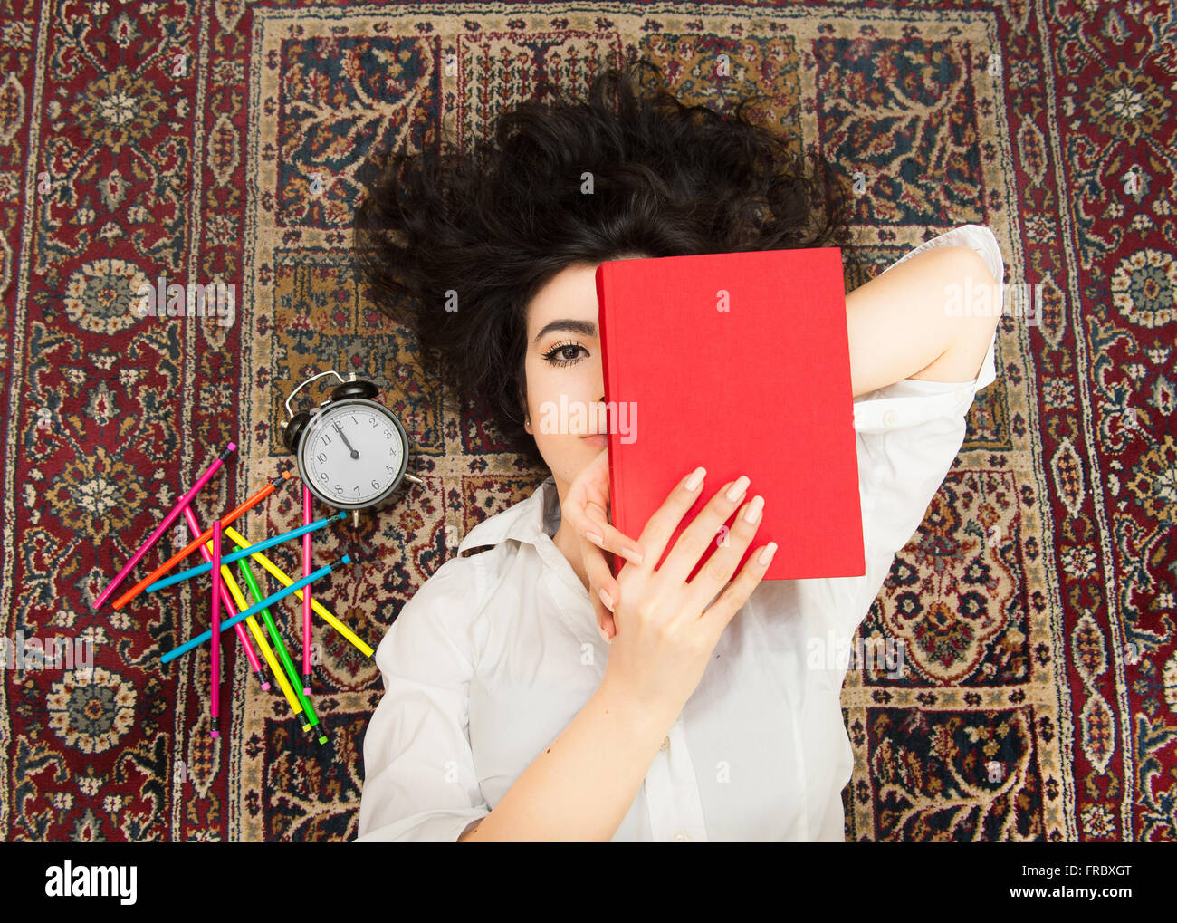 Young woman student lying on carpet with red book clock and pencils ...