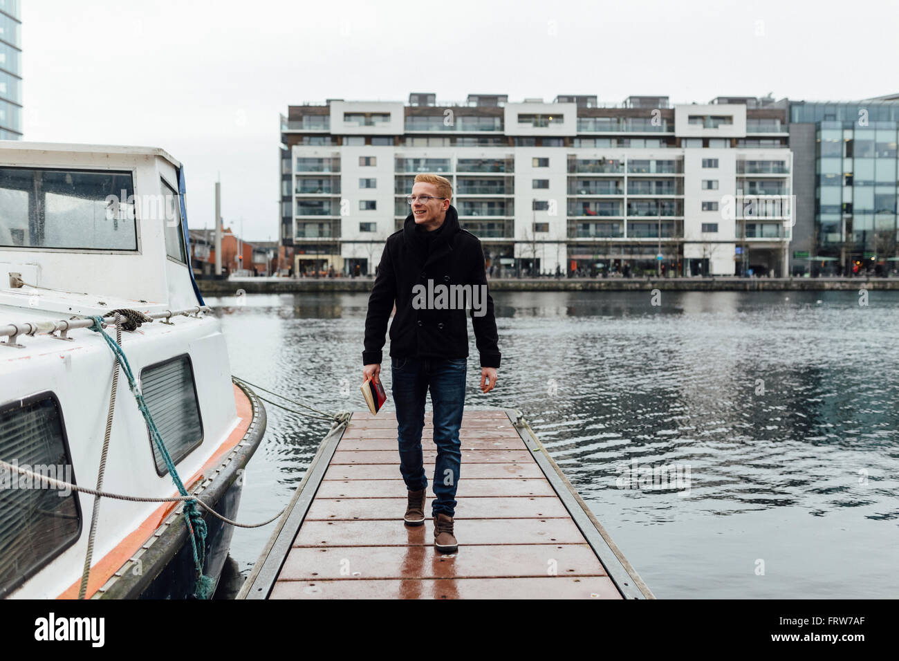 Ireland, Dublin, smiling young man with a book walking on a jetty in ...