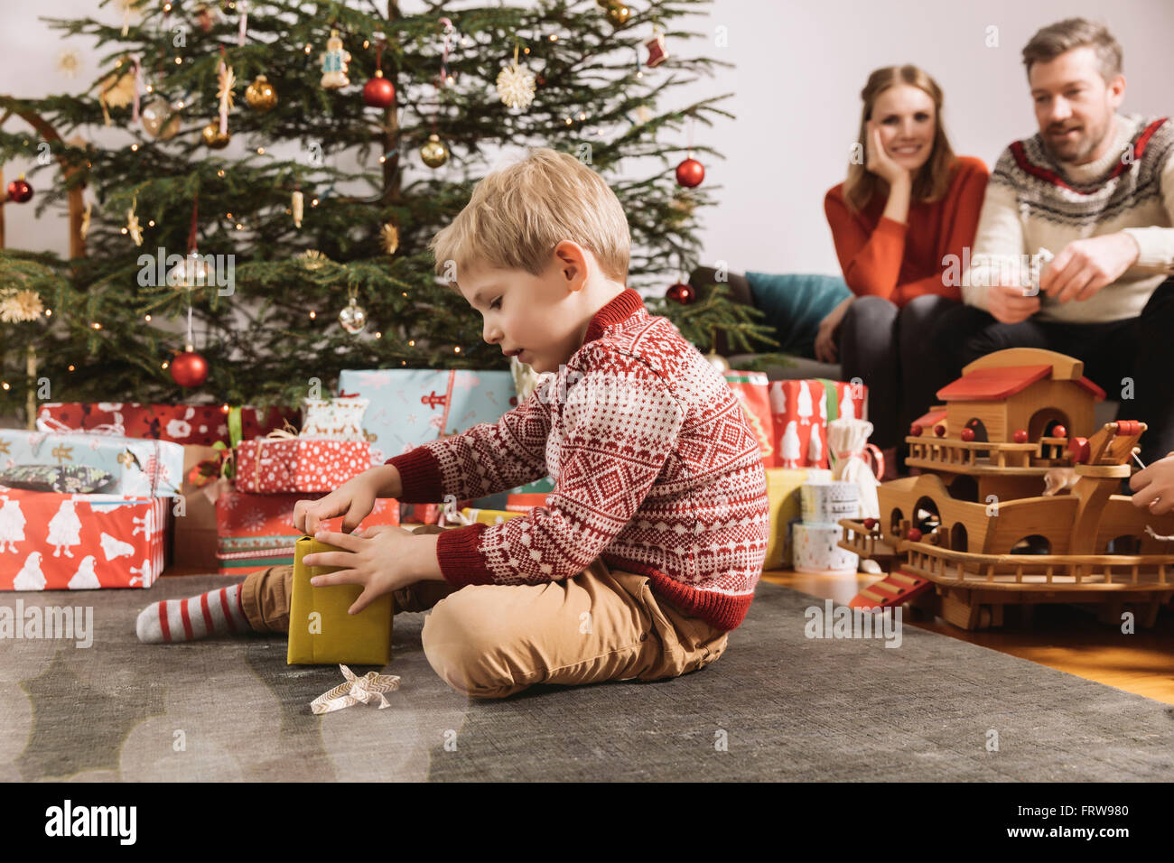 Little boy opening Christmas gift with parents watching in background ...