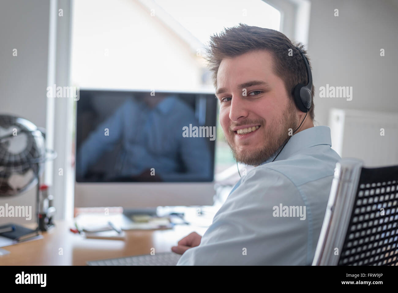 Portrait of smiling young man at desk in office wearing headphones ...