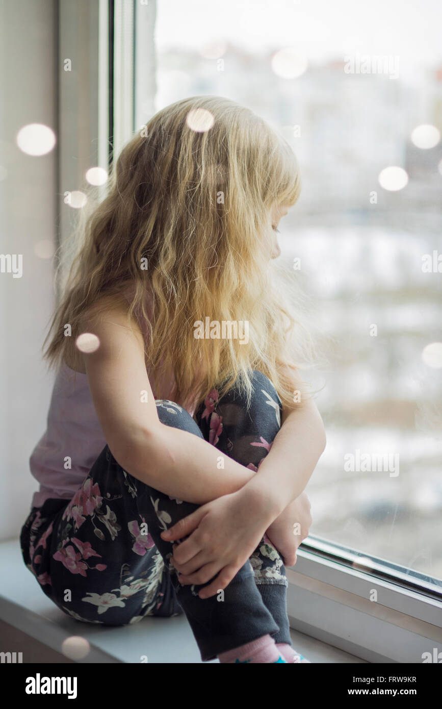 Blond little girl sitting on window sill looking through window Stock ...