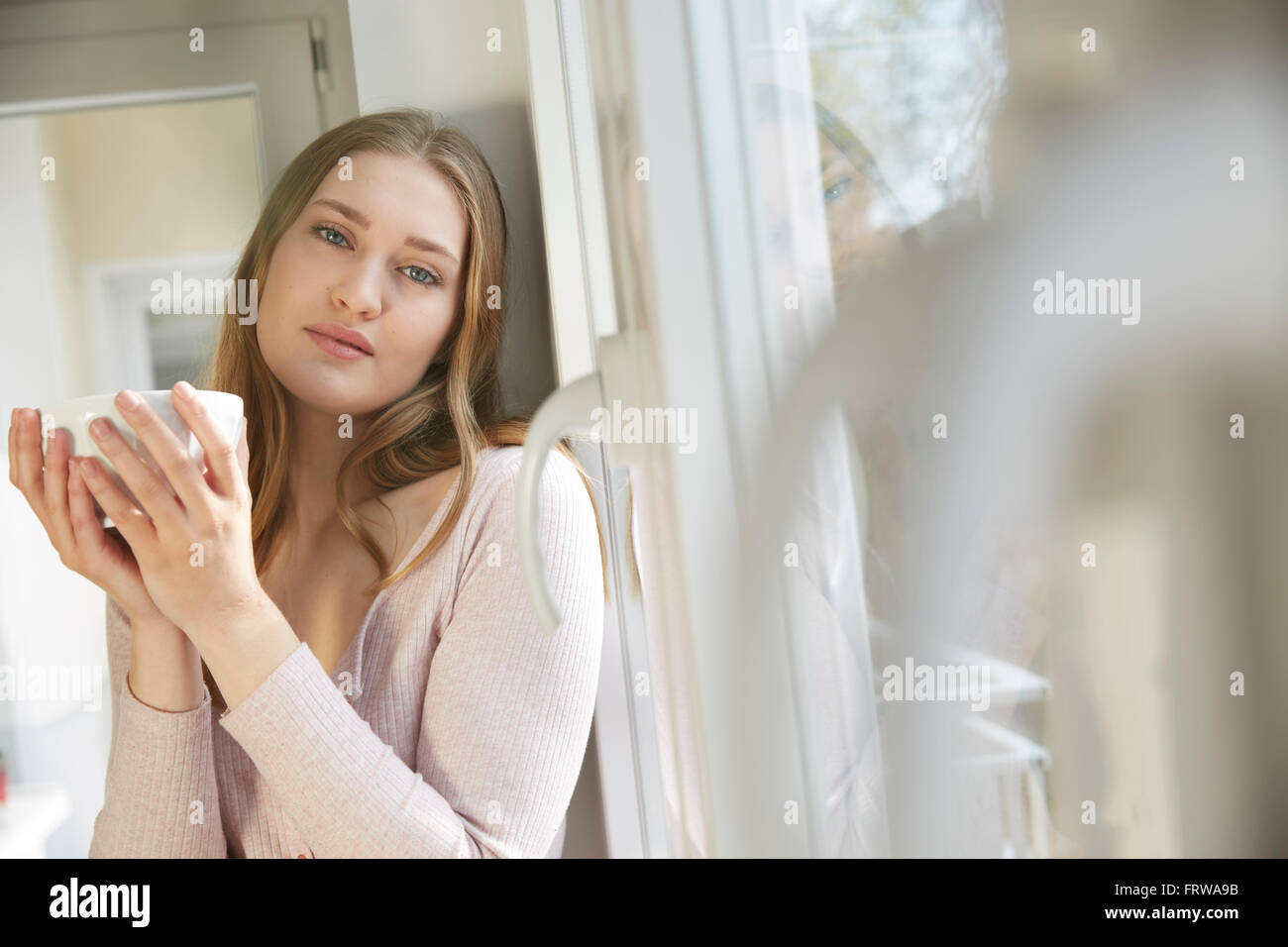 Portrait of blond woman with bowl of coffee leaning against window ...