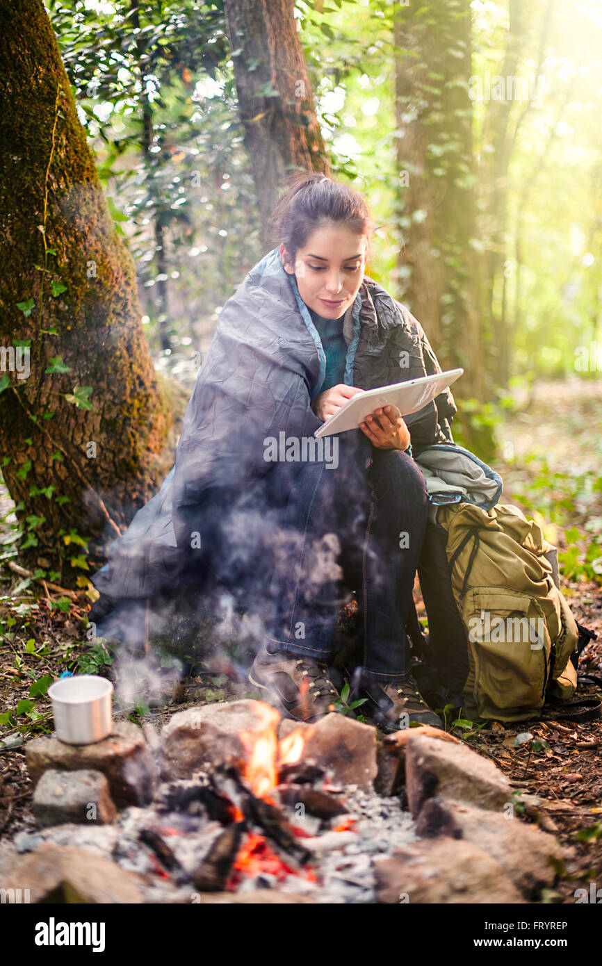 in the woods a young woman sitting near a campfire using a digital ...