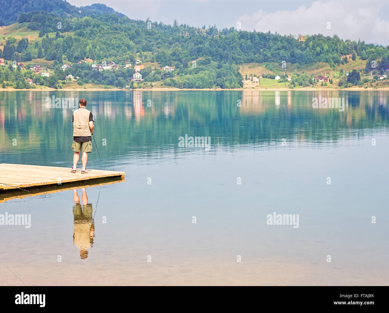 Young man in shorts and vest fishing standing on a floating wooden pier ...