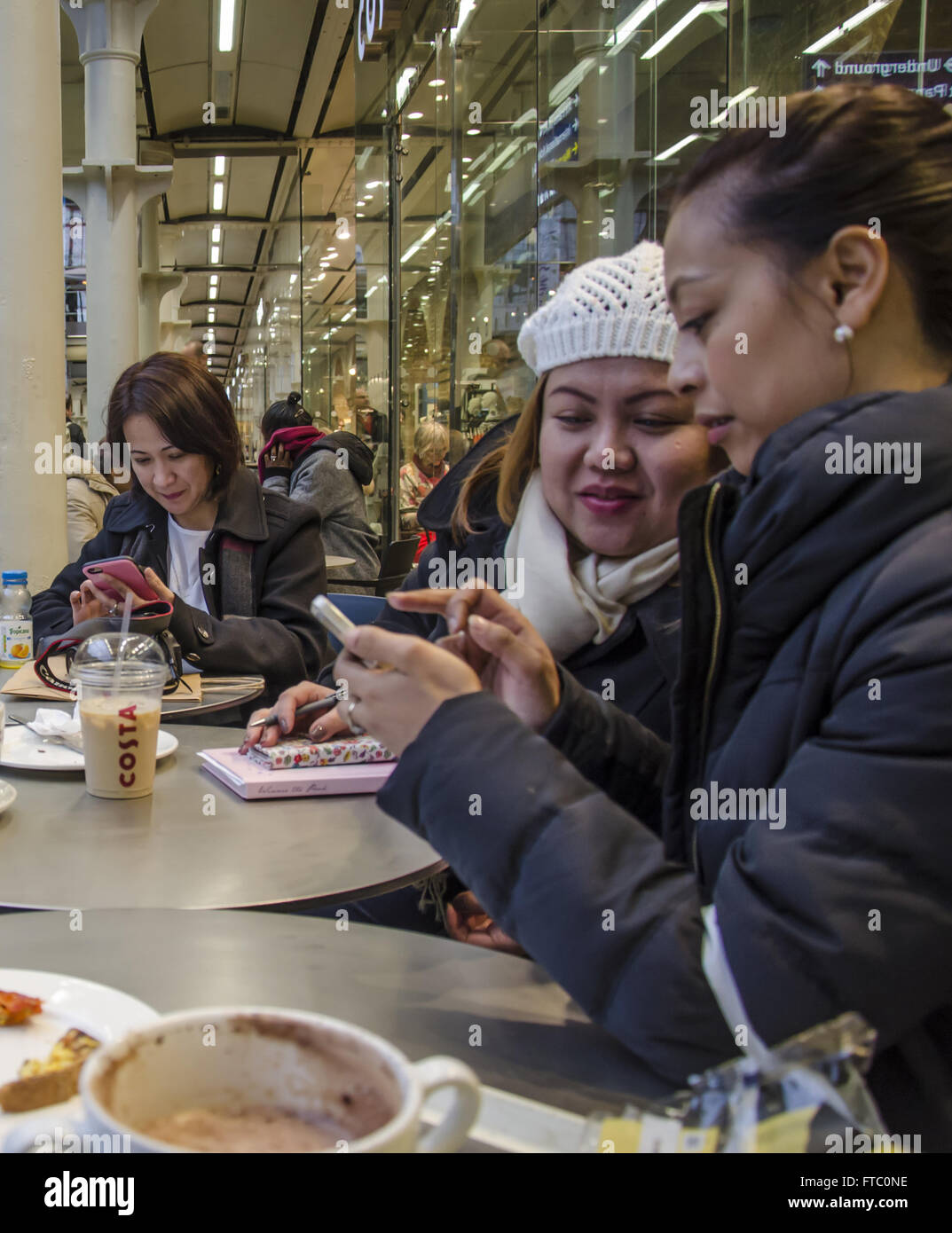 Friends sit together at tables outside a cafe but are all busy sending ...