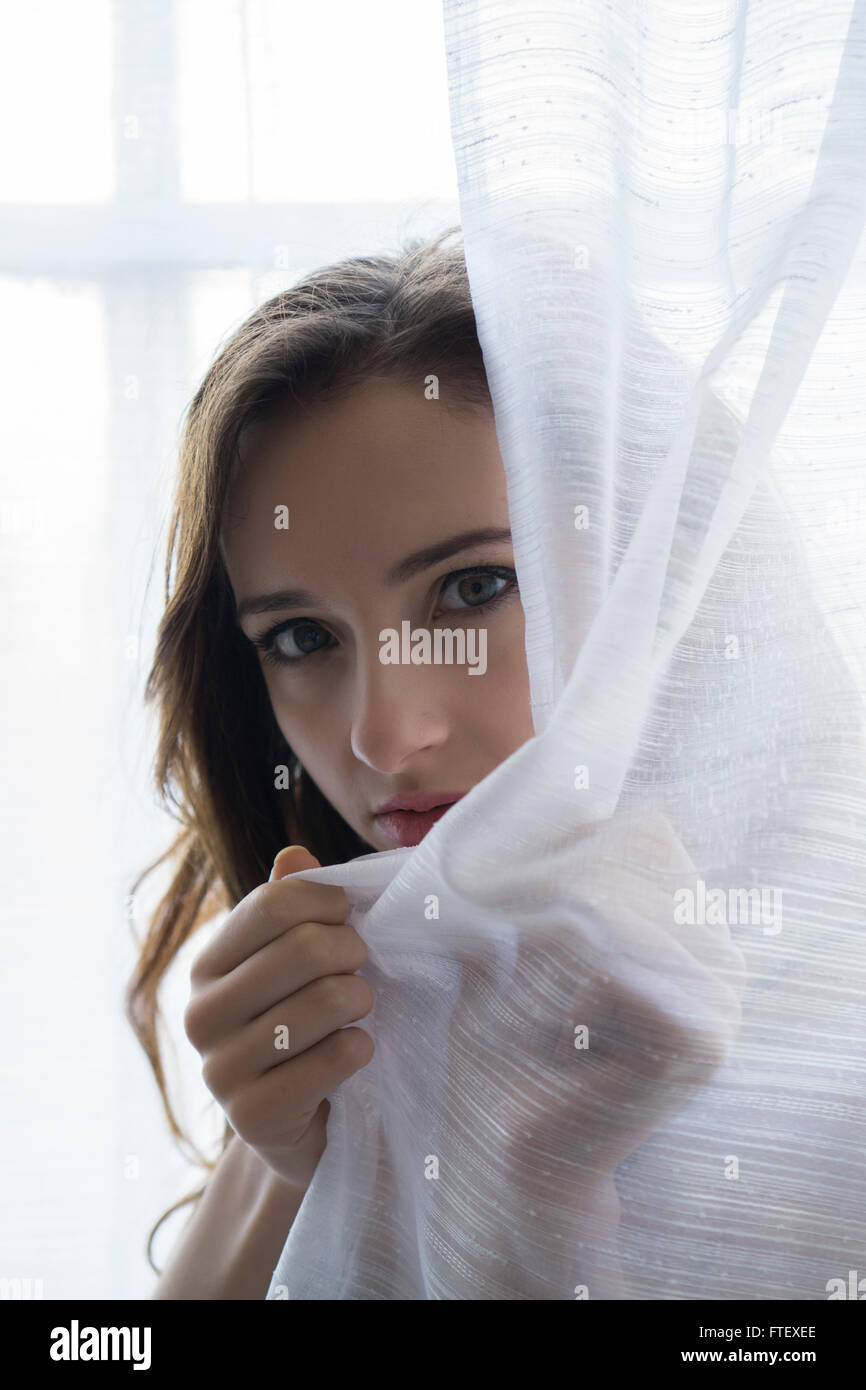 Serious young woman hiding behind the curtains by the window Stock ...