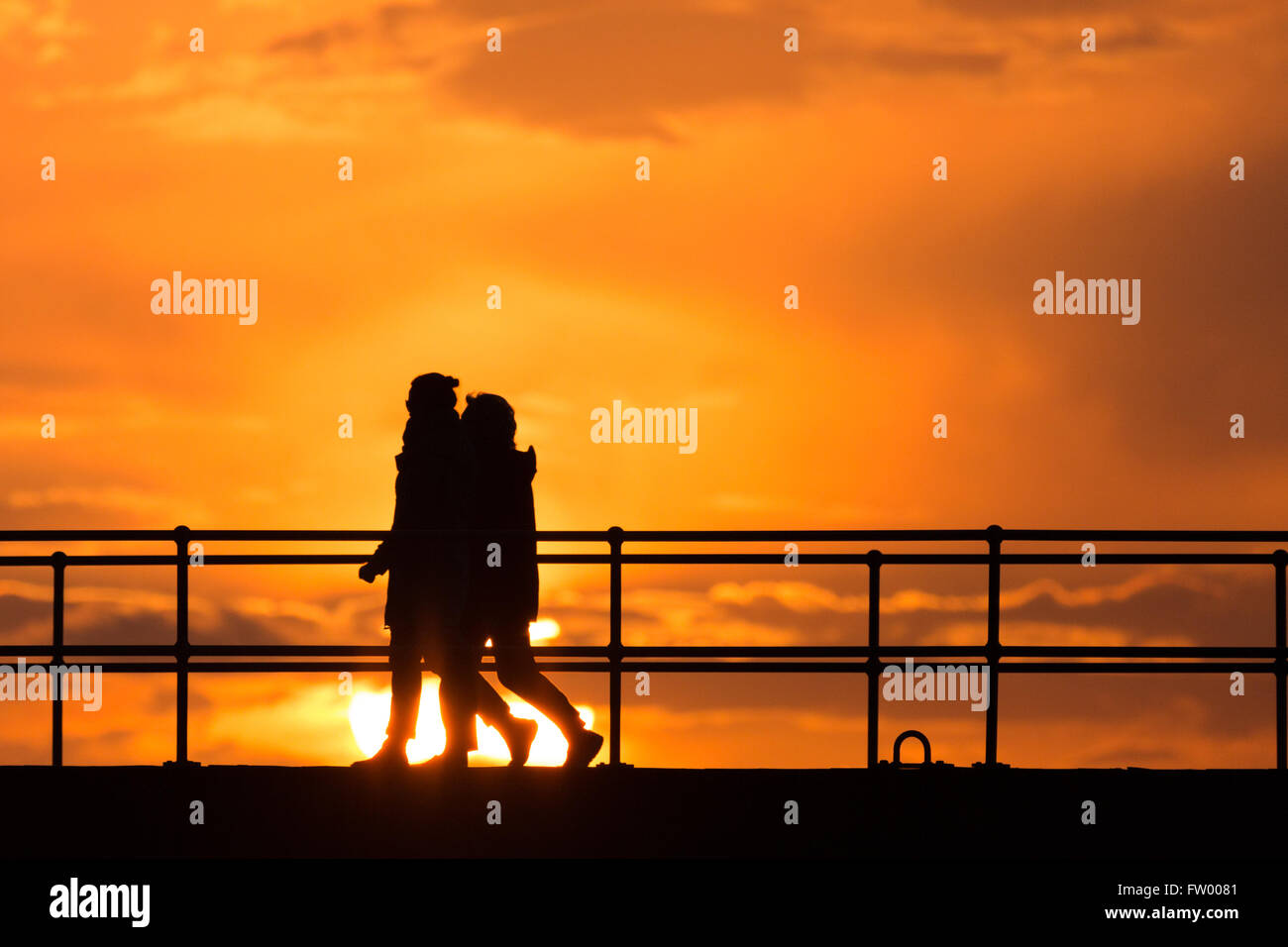 Two people silhouetted as they walk along a railed jetty at sunset ...