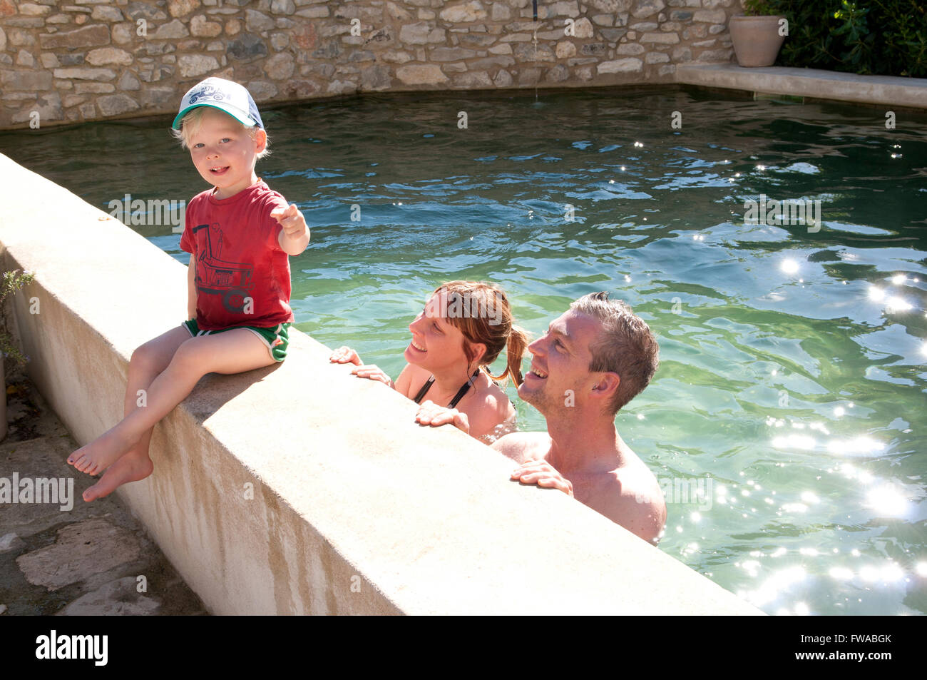 Toddler sitting on the edge of a swimming pool with his parents ...