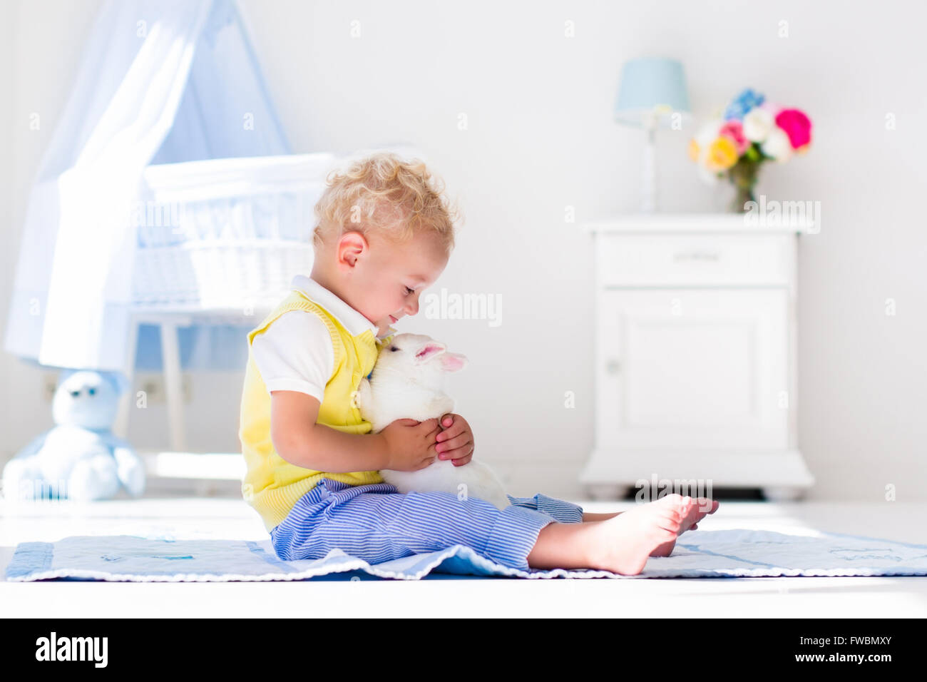 Cute blond curly baby boy playing with rabbit in a white sunny bedroom ...