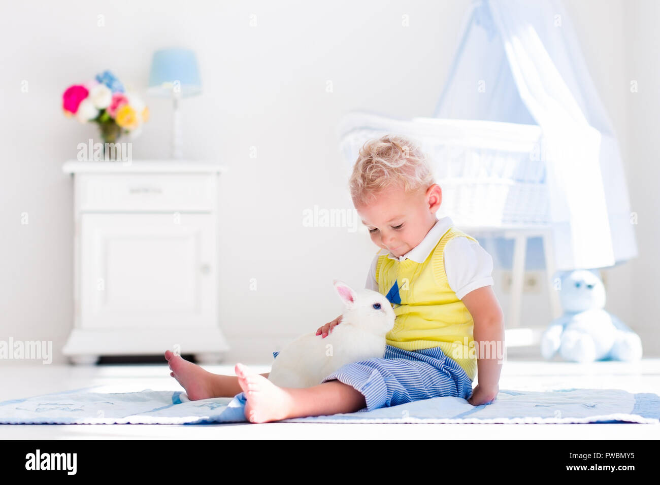 Cute blond curly baby boy playing with rabbit in a white sunny bedroom ...