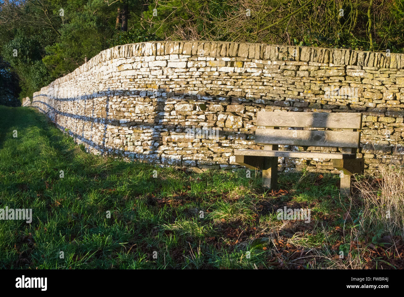A long dry stone wall in the Cotswolds, Uk with an empty bench next to ...