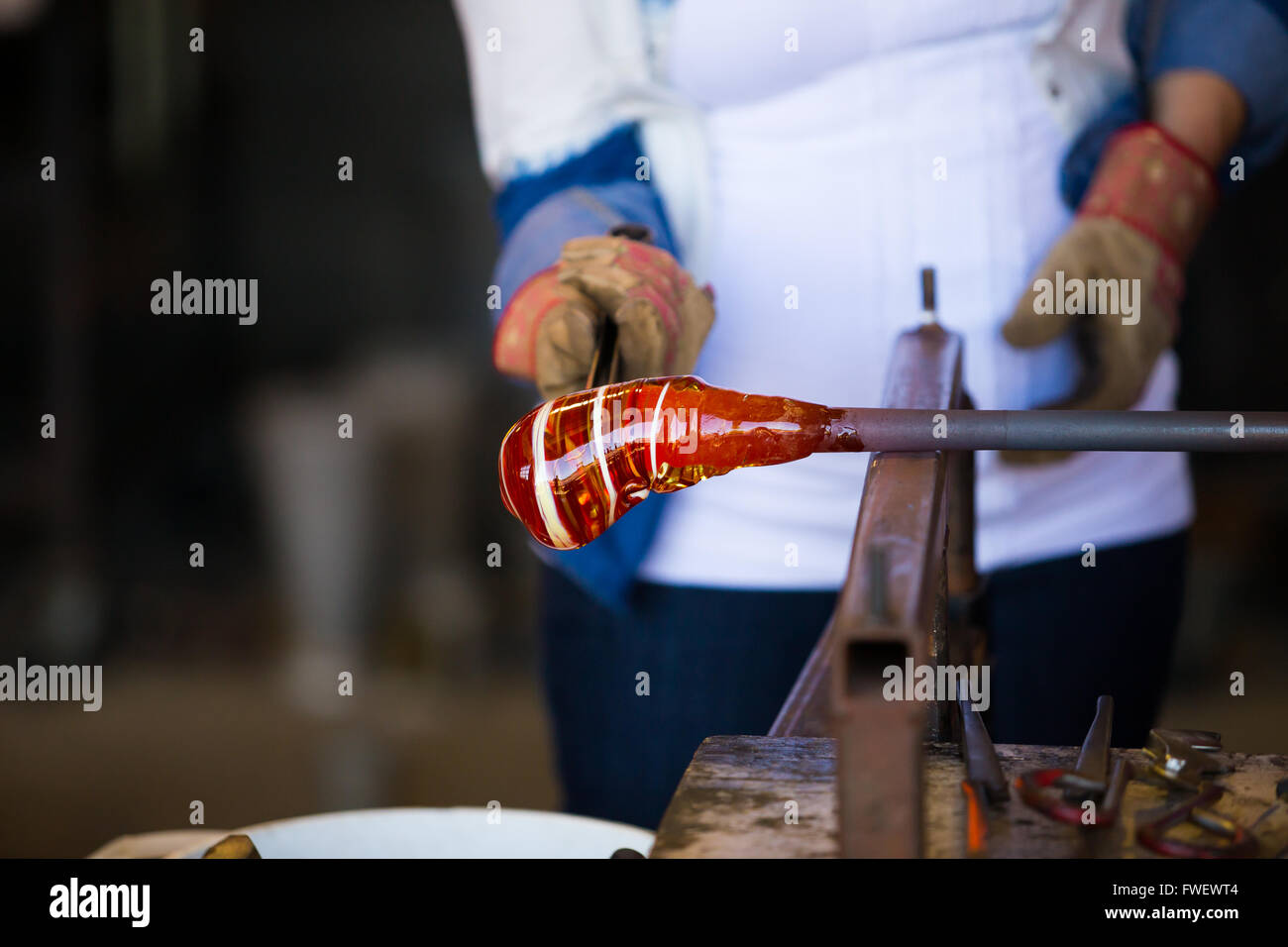 A female is blowing glass and using a wooden shaping tool to form the ...
