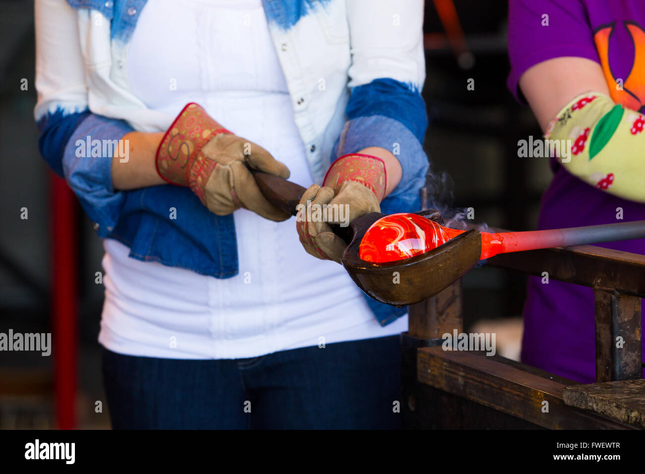 A female is blowing glass and using a wooden shaping tool to form the ...