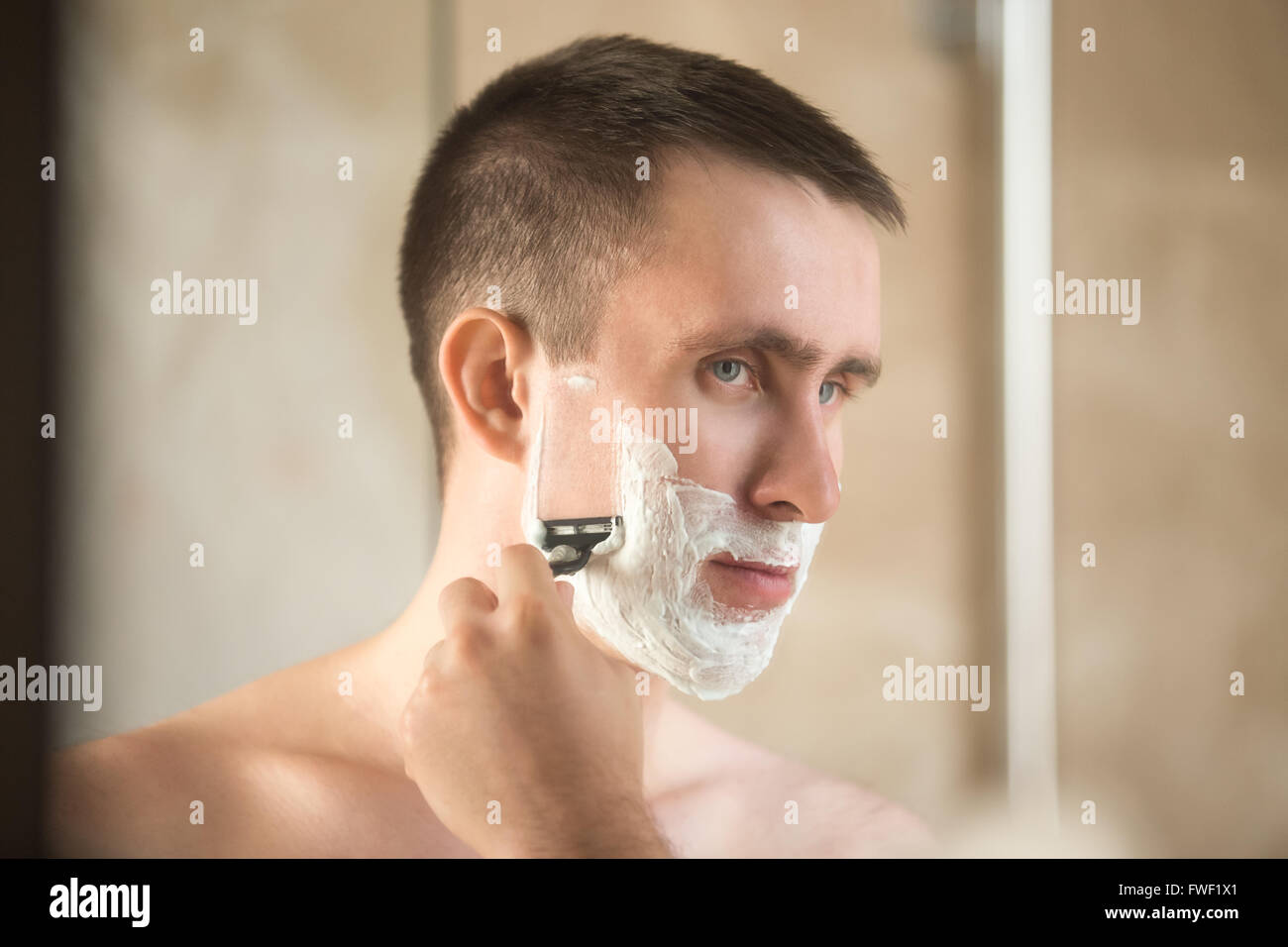 Portrait of handsome young man shaving his face in the bathroom in ...