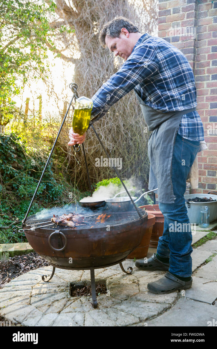 A man cooking roasting game birds over an open fire, pouring olive oil ...