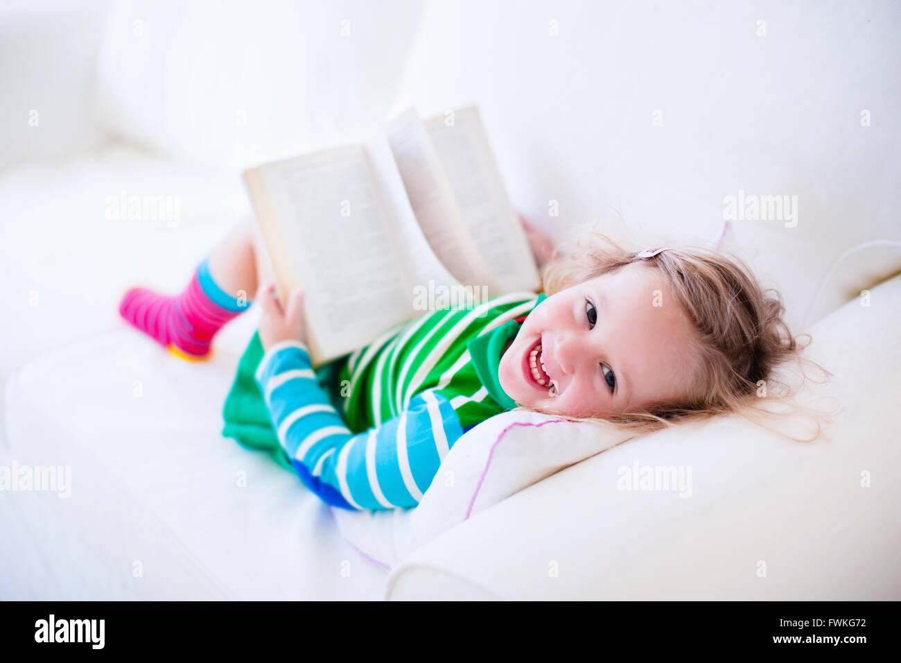 Little girl reading a book relaxing on a white couch. Kids read books ...