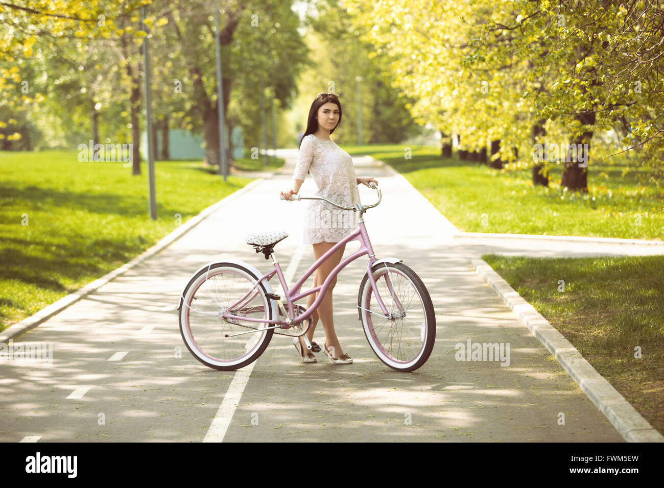 Young brunette girl in sundress riding a bicycle in green park at ...