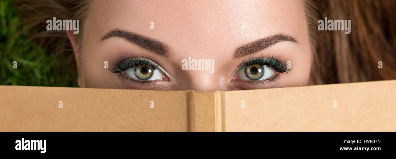 Beautiful young woman looking over book cover laying on grass ...