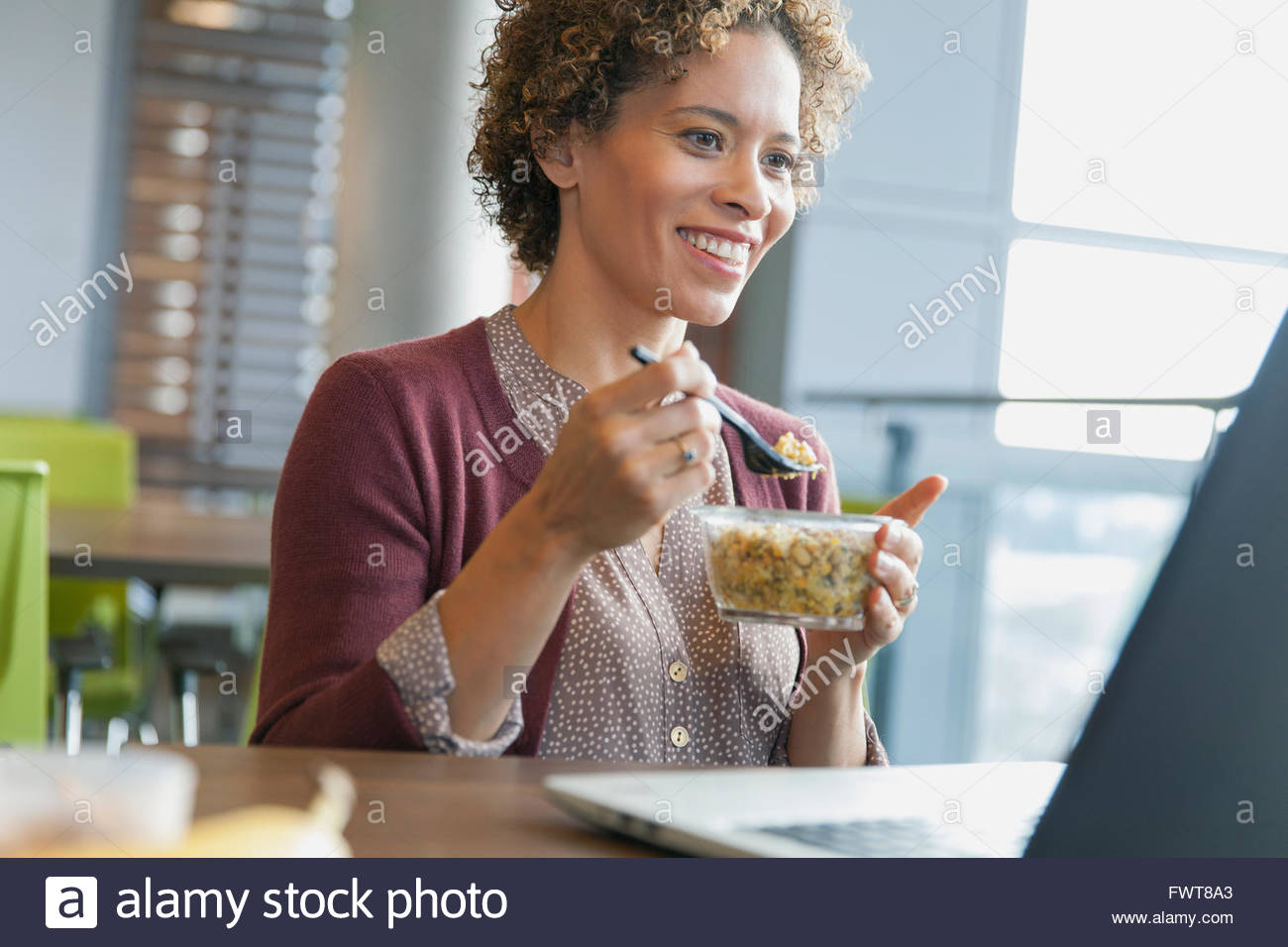 Woman smiling as she snacks at her laptop Stock Photo - Alamy