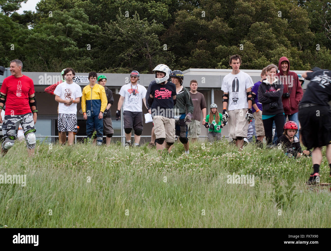 Skateboarders (mostly young men) stand on a grass bank watching ...