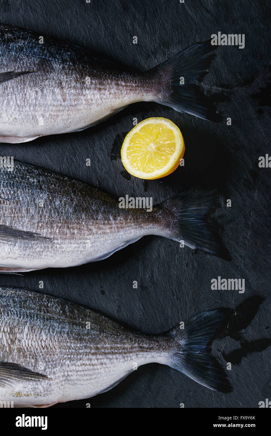 Three tails of raw bream fish with lemon on stone slate black textured ...