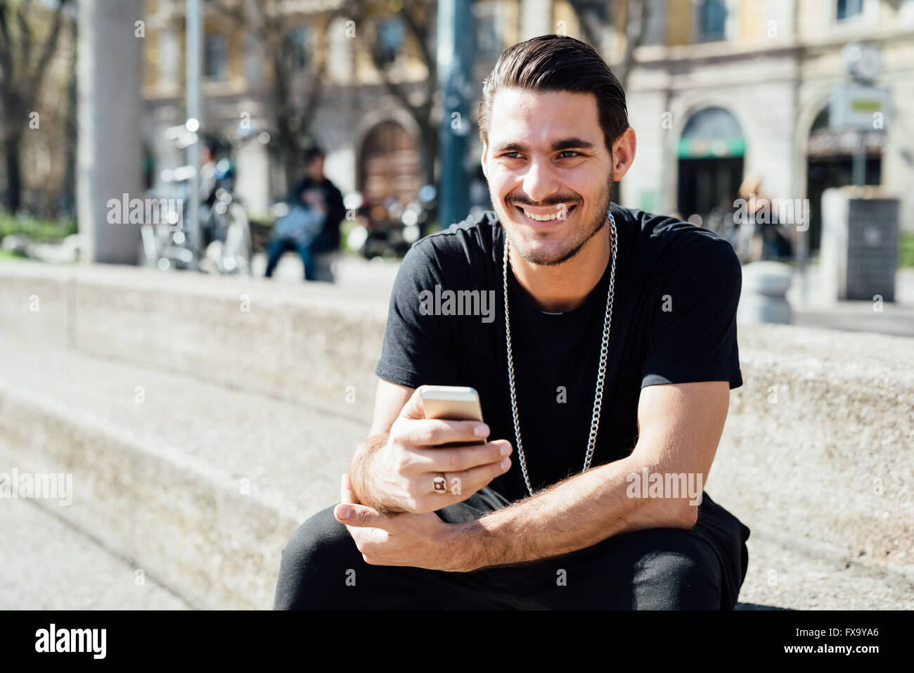Young handsome italian boy seated on a sidewalk using a smart phone ...