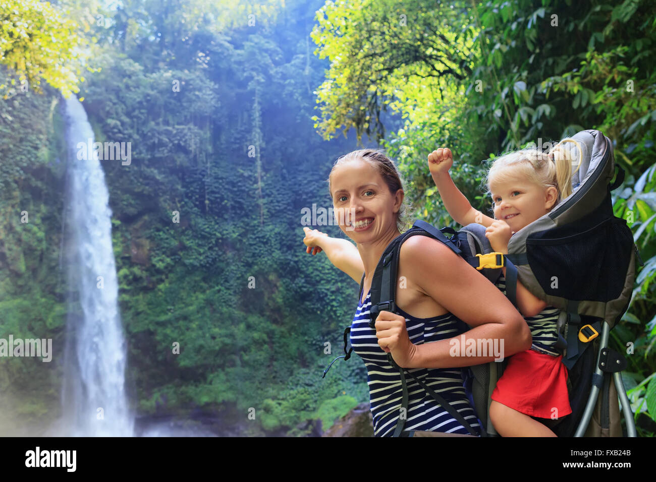 Mother hold baby girl on back in carrying backpack walking in waterfall ...